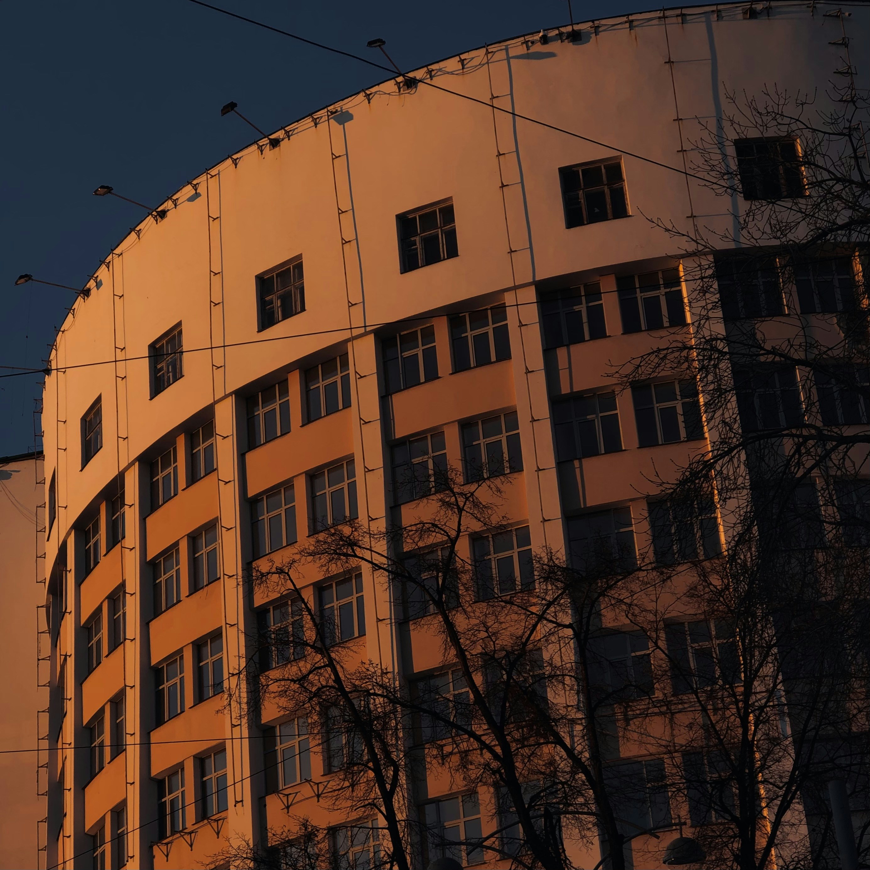 Curved modern building illuminated by warm sunset light, with silhouetted trees in the foreground.