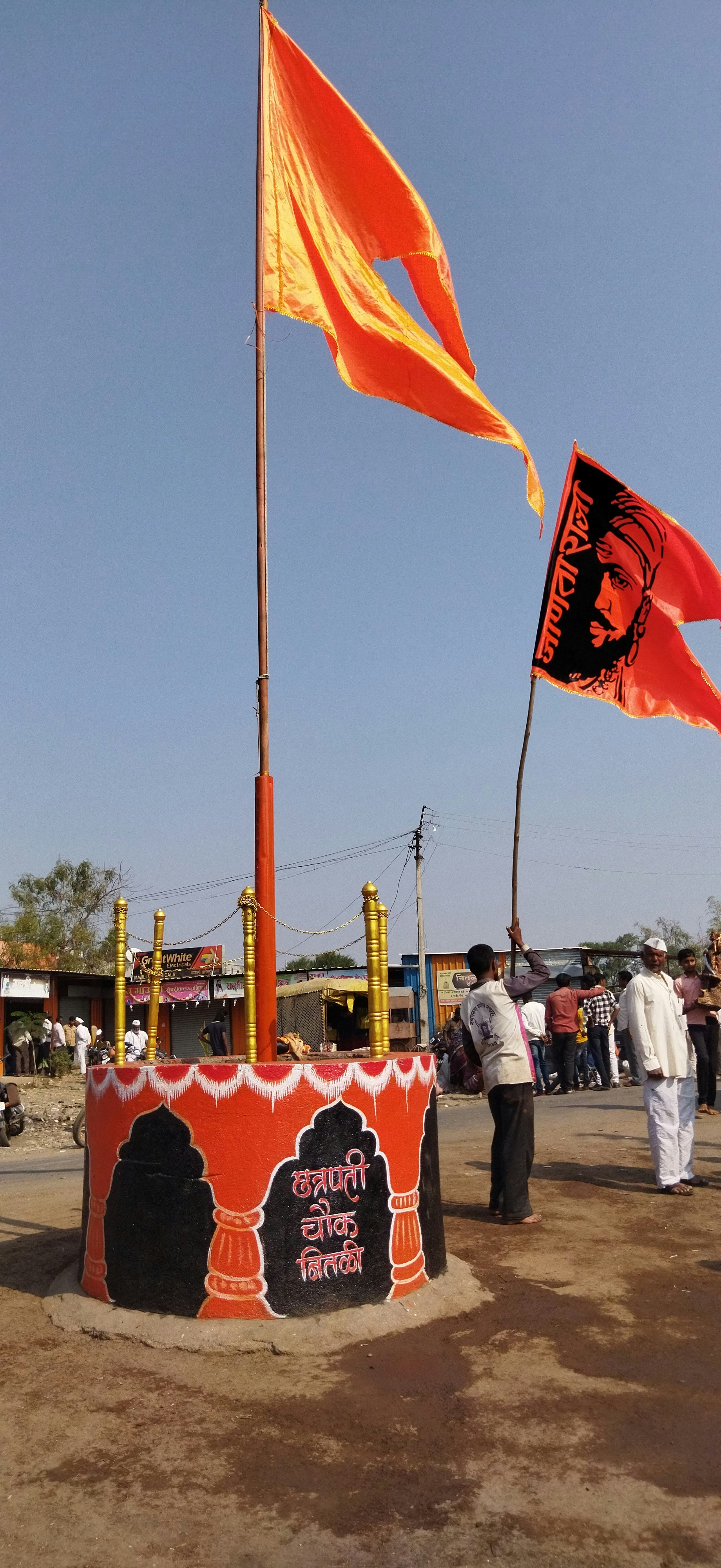 A colorful flagpole adorned with orange flags stands prominently in a bustling outdoor setting, surrounded by people celebrating a local festival.