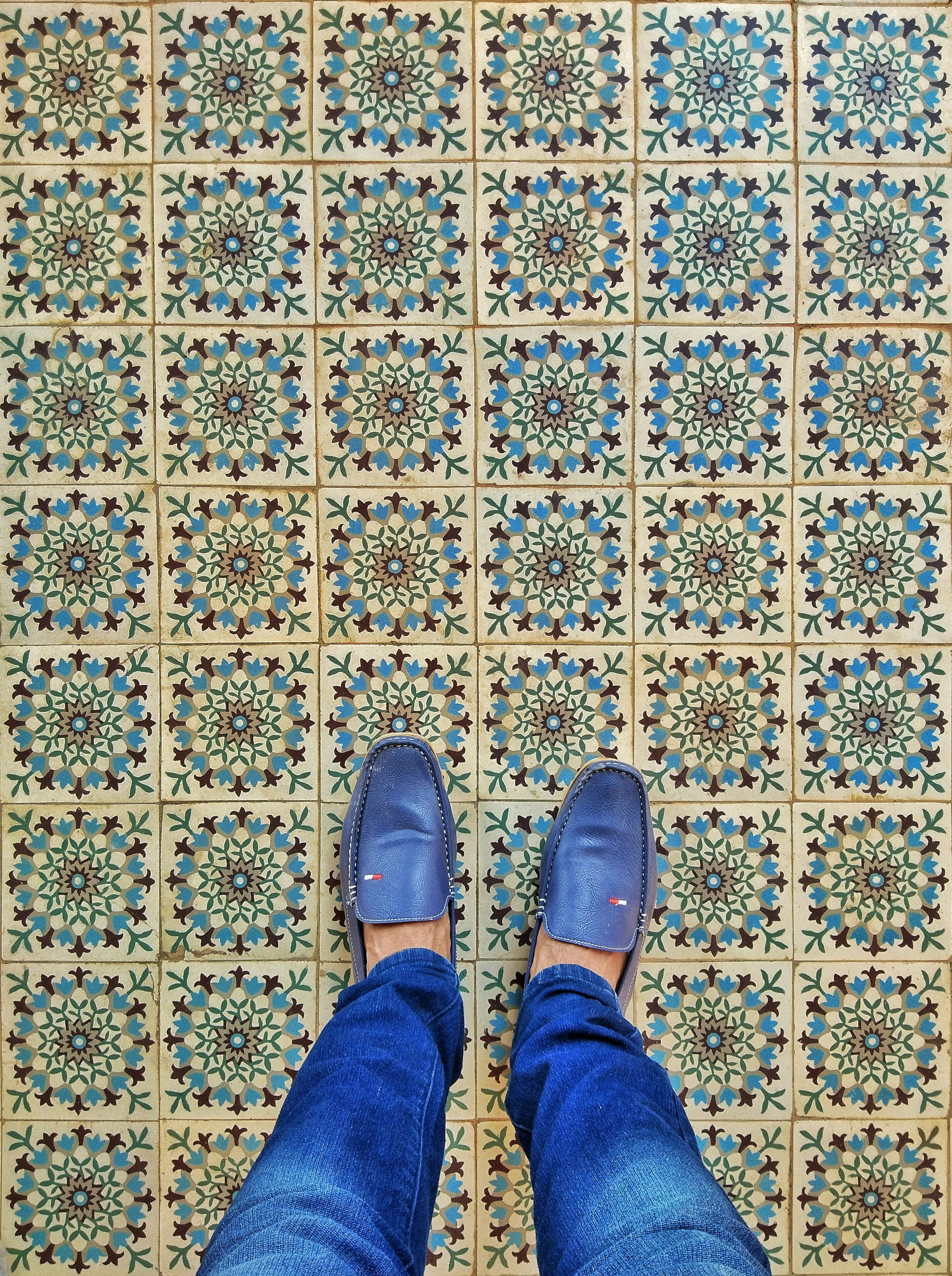 Intricate patterned tiles create a vibrant backdrop, with a pair of blue shoes standing prominently at the center. The design showcases a blend of floral motifs and geometric shapes.