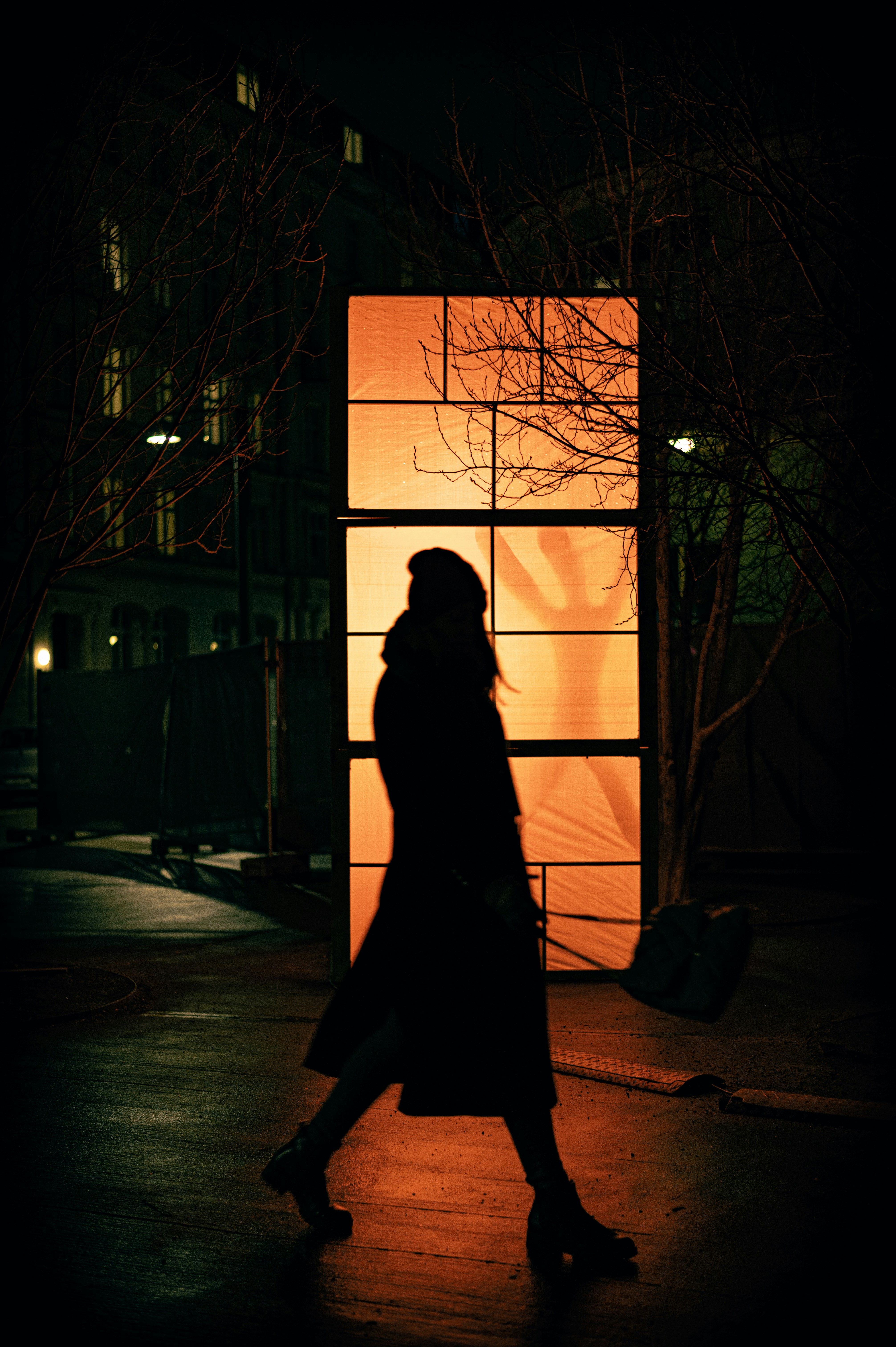 A silhouette of a woman walking past a glowing backdrop with an abstract handprint, creating a mysterious urban scene at night.