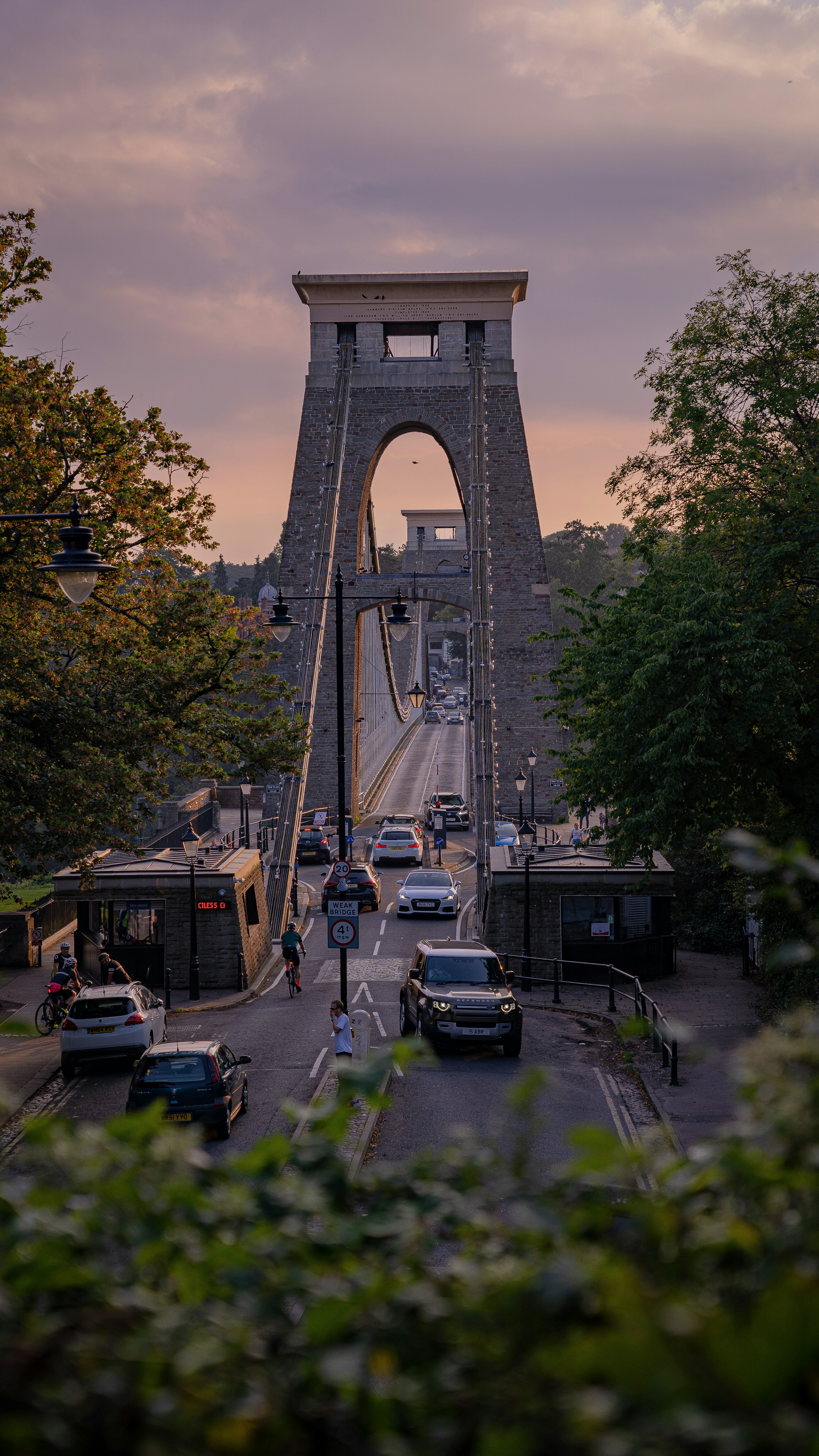 Clifton Suspension Bridge spanning a busy road, framed by lush greenery and a pastel sky at dusk.