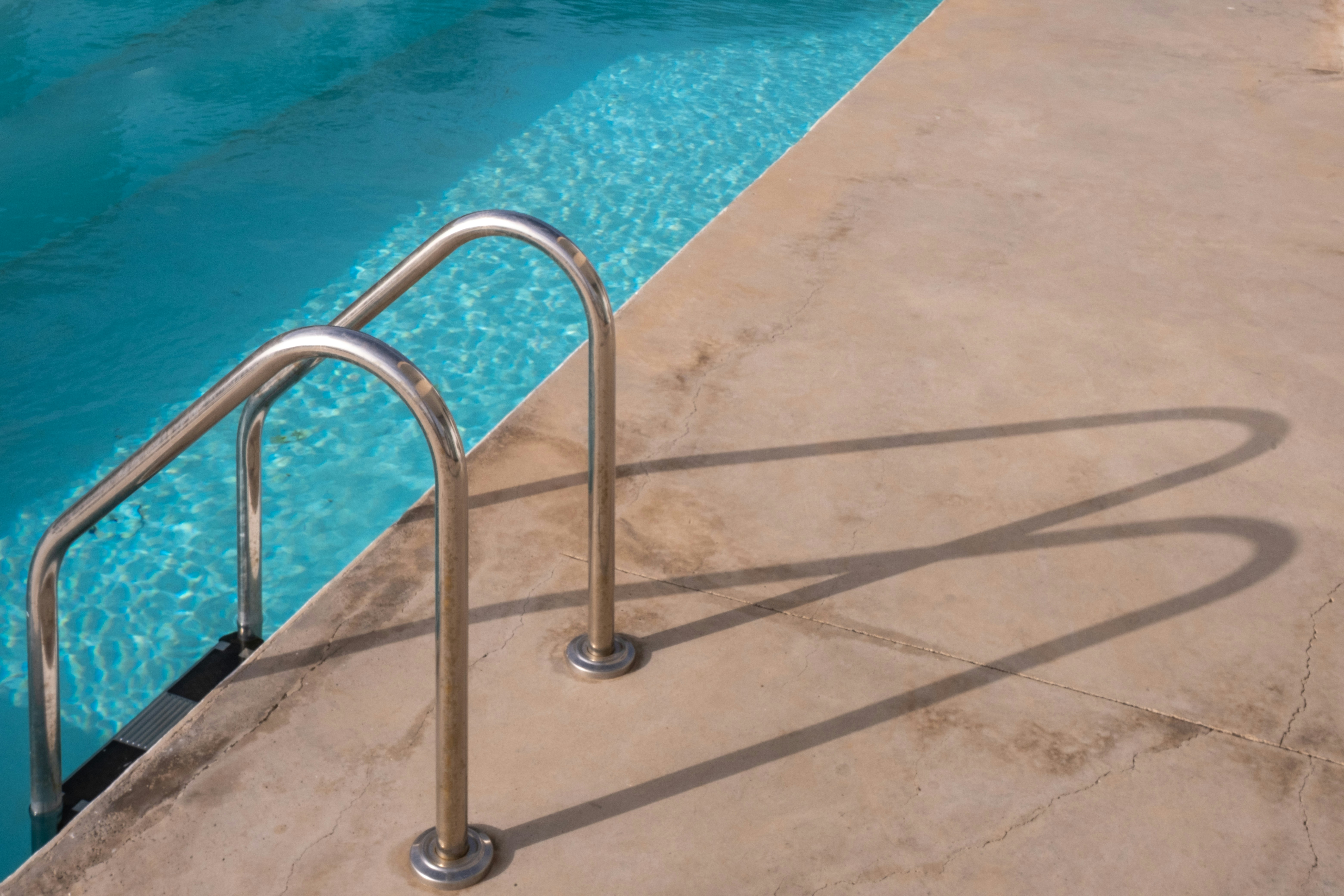 Stainless steel pool ladder casting intricate shadows on a sunlit concrete surface beside a sparkling blue pool.