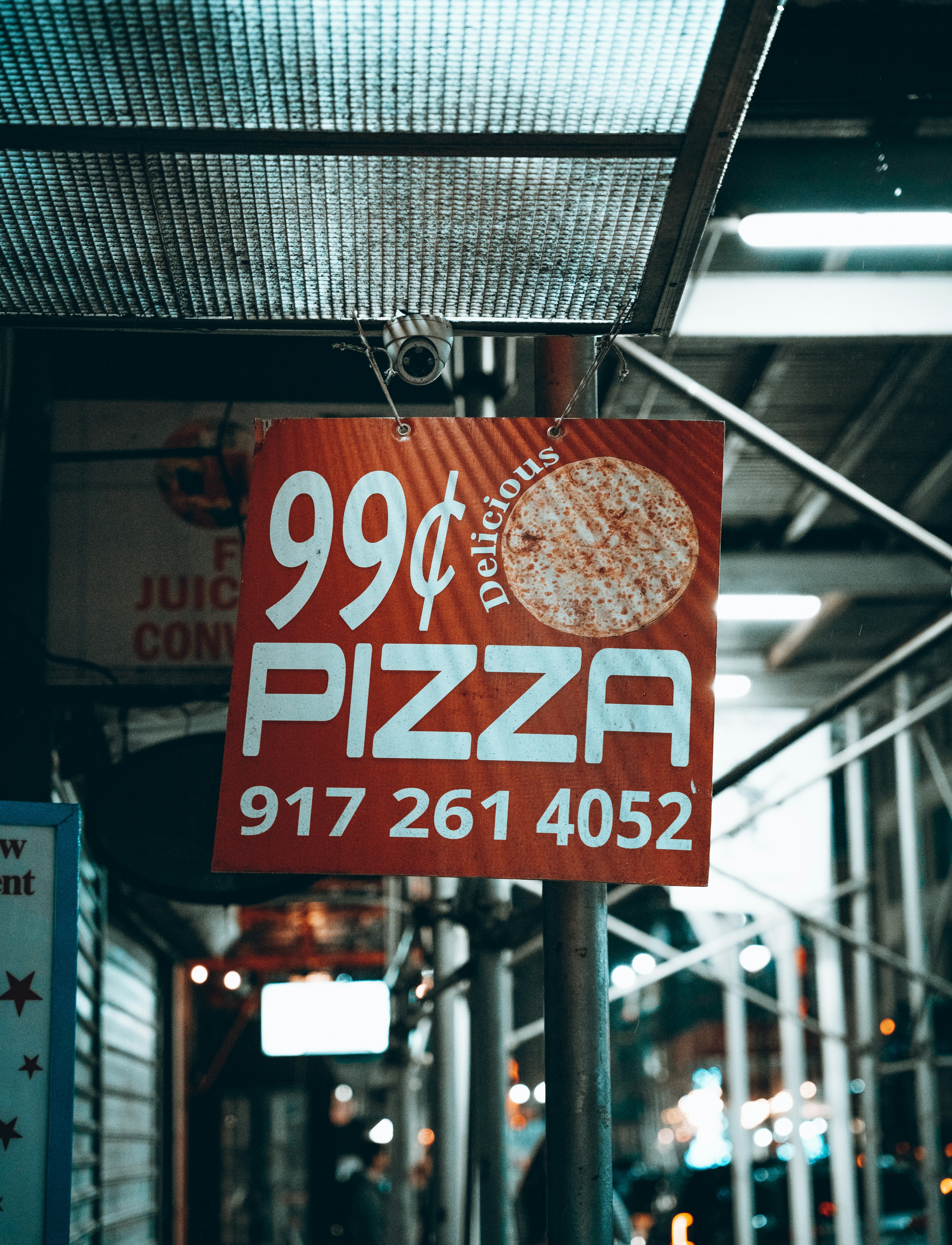 Bright red sign advertising 99 cent pizza, featuring bold white text and a pizza graphic. The sign hangs in an urban setting, reflecting the hustle and bustle of city life.