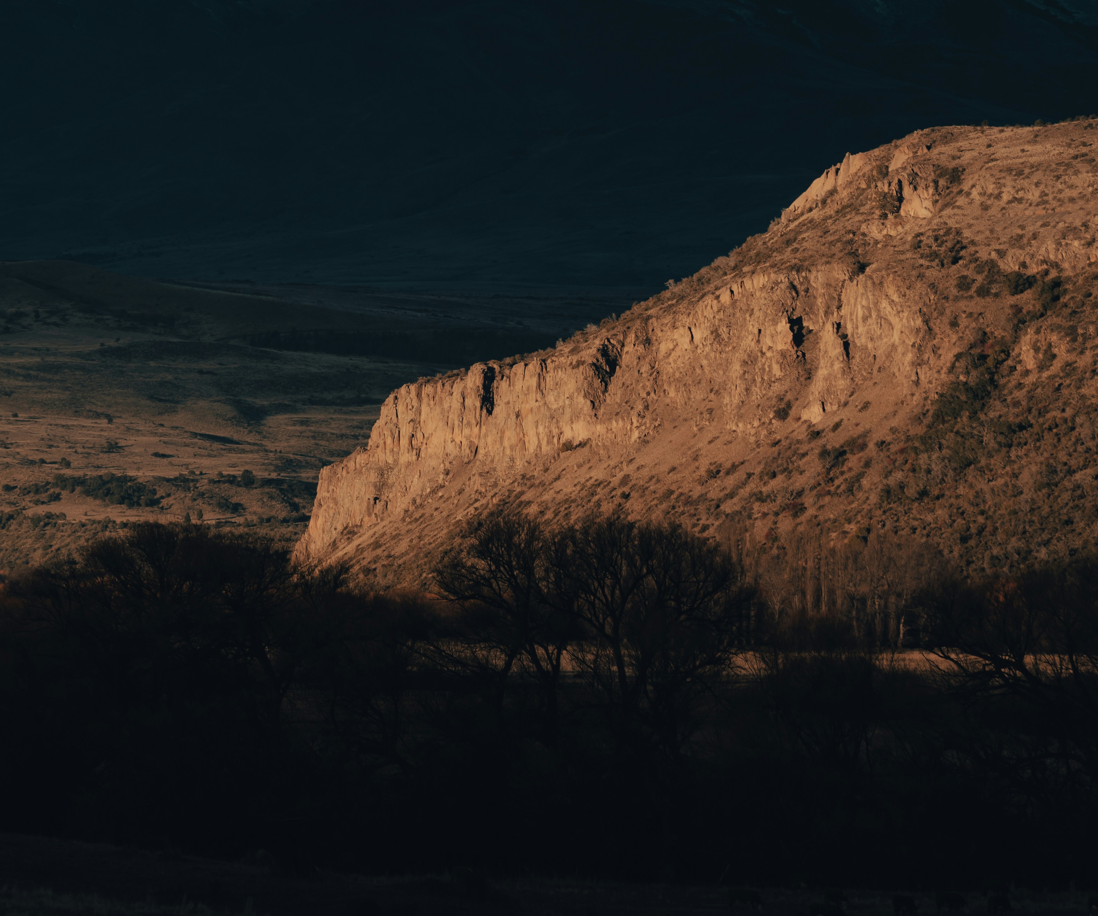 a mountain side with trees in the foreground and a dark sky in the background
