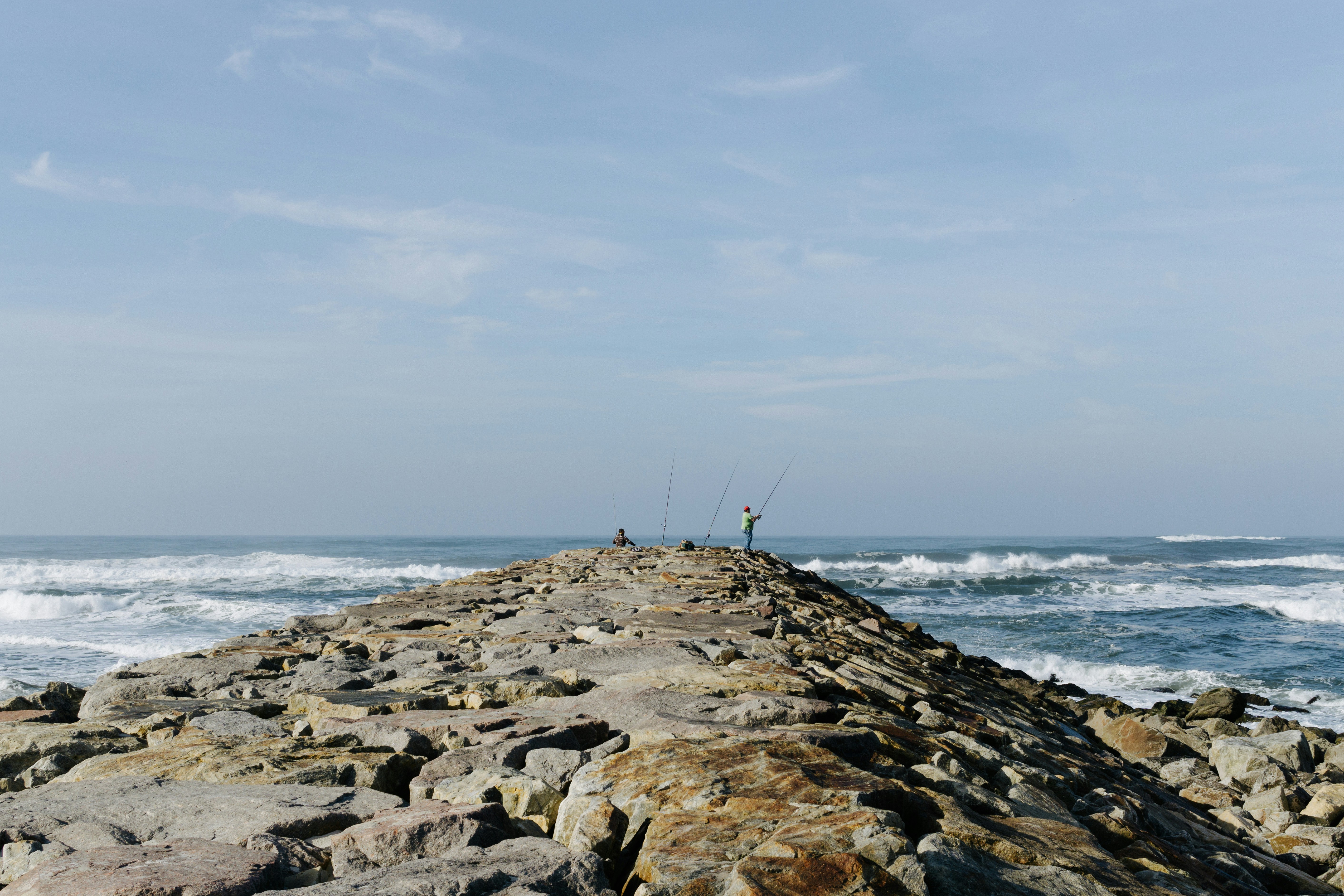Two fishermen casting lines from a rocky jetty, surrounded by crashing waves under a clear sky.
