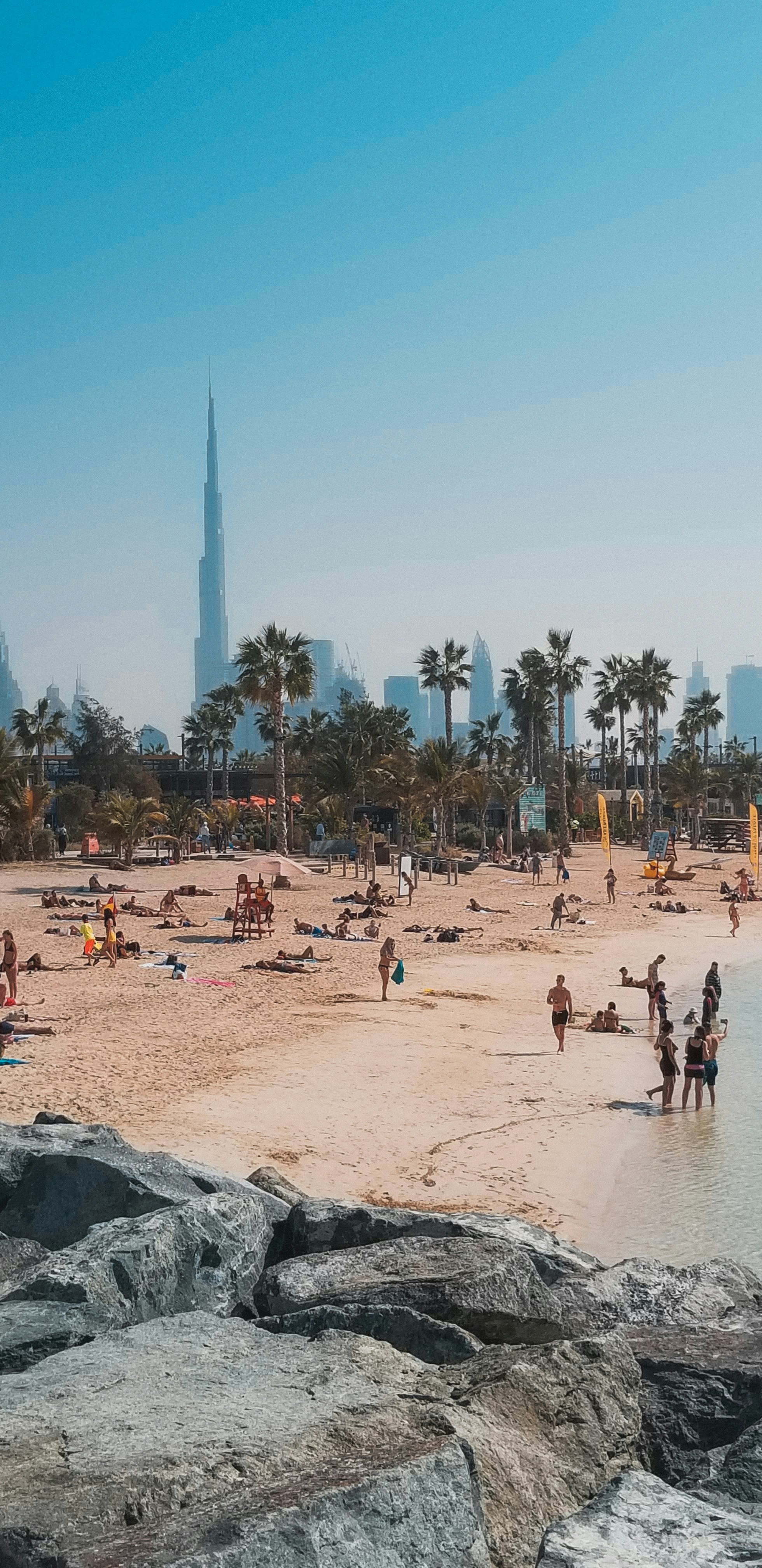Vibrant beach scene with sunbathers and palm trees, set against the backdrop of towering skyscrapers, including the iconic Burj Khalifa. A perfect blend of relaxation and urban life.