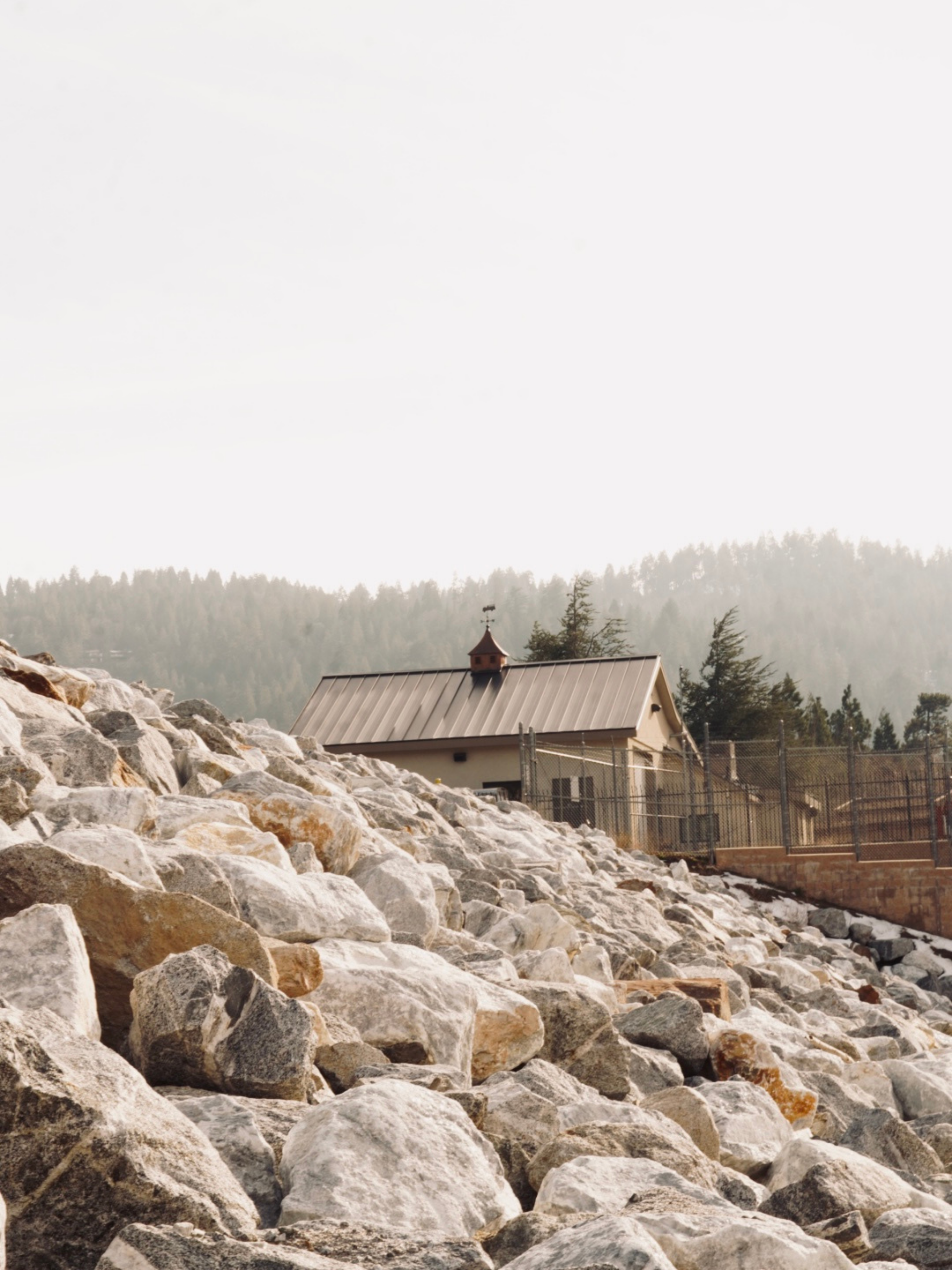 Rocky shoreline leading to a quaint building nestled among trees, with a soft fog enveloping the background.