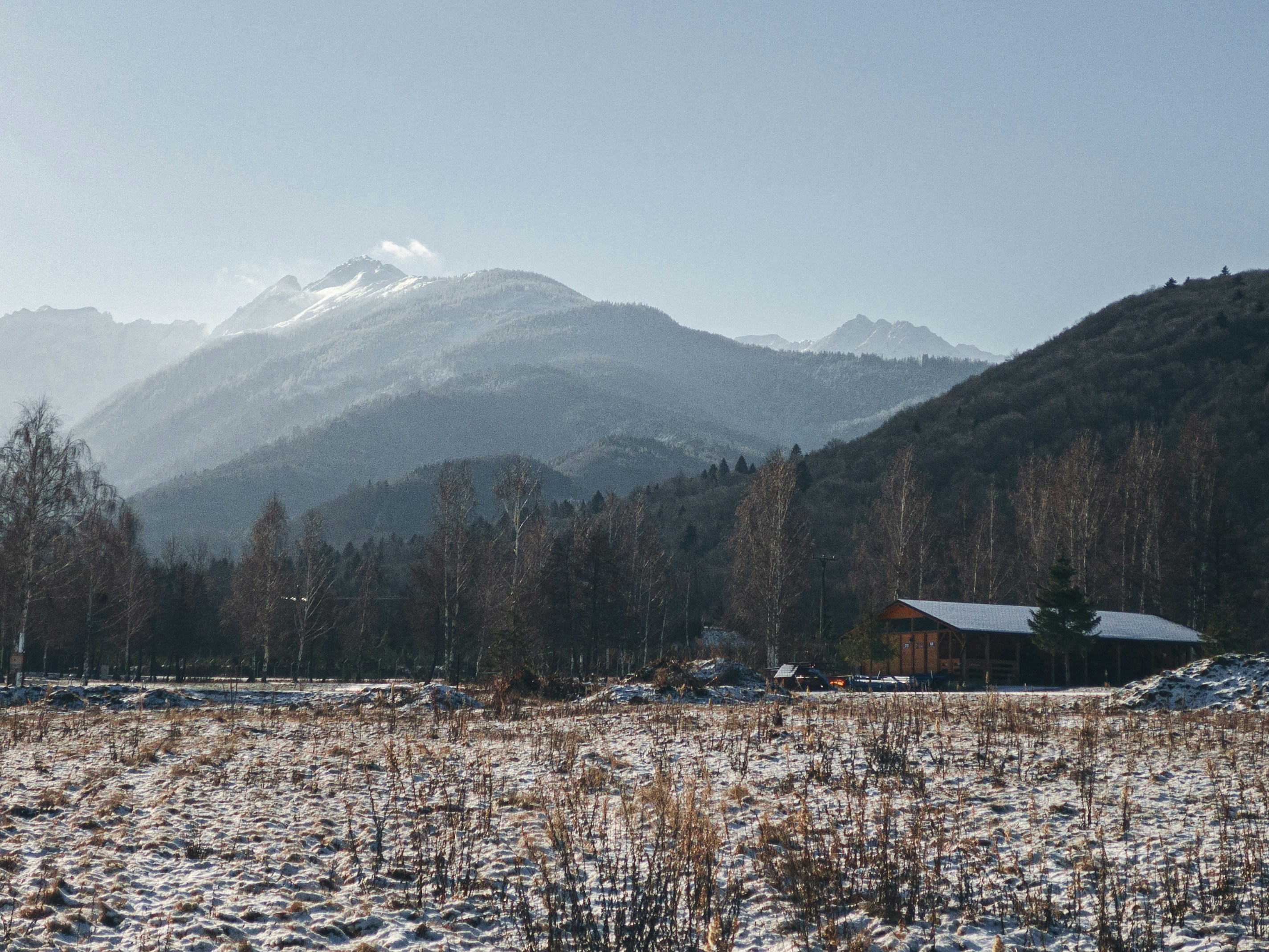 Snow-covered mountains loom over a tranquil landscape, with a rustic cabin nestled among the trees. The scene captures the serene beauty of winter's embrace.