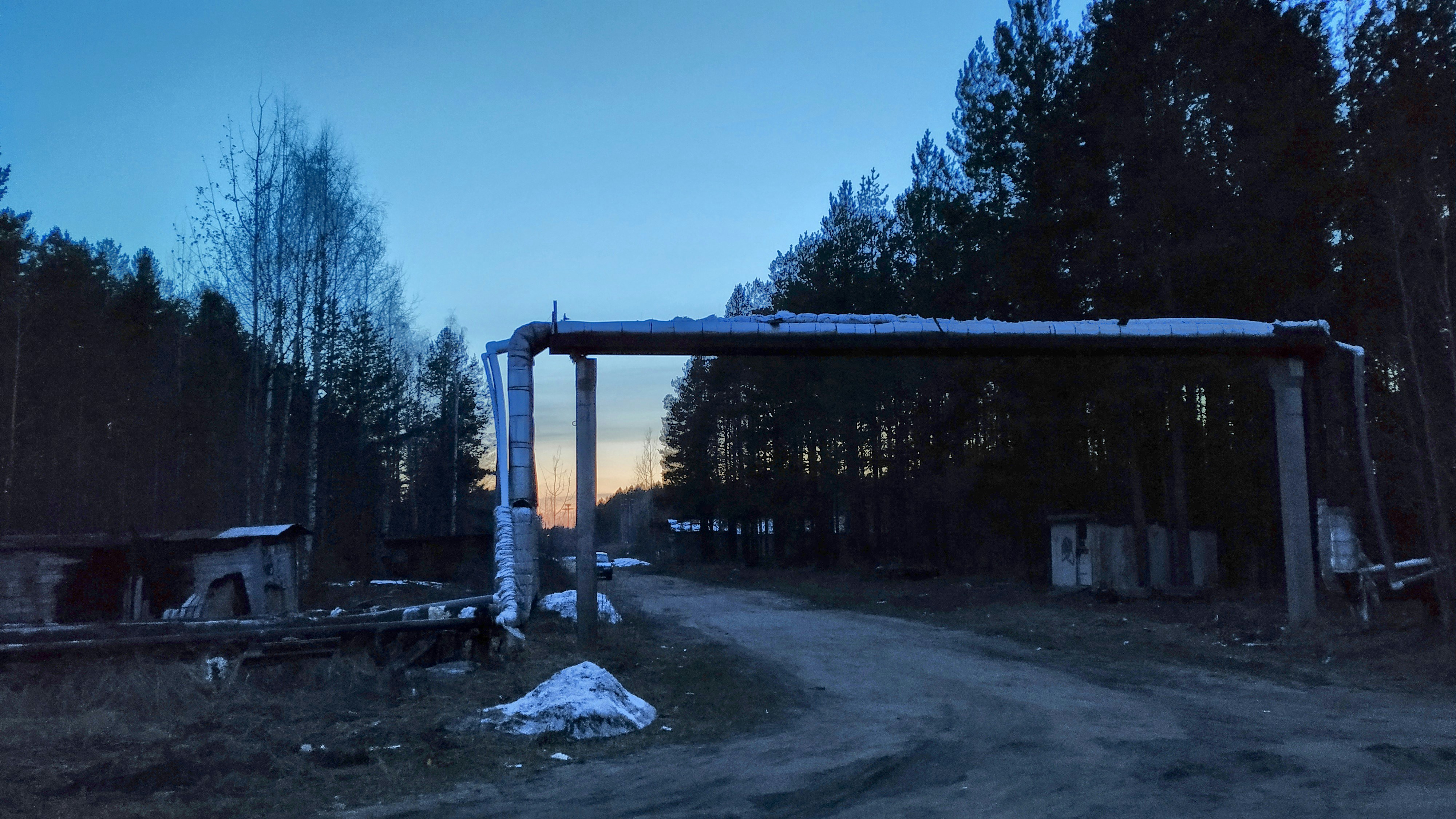 An abandoned industrial site at dusk, featuring a dilapidated archway and remnants of structures, surrounded by trees. The fading light casts a haunting atmosphere.