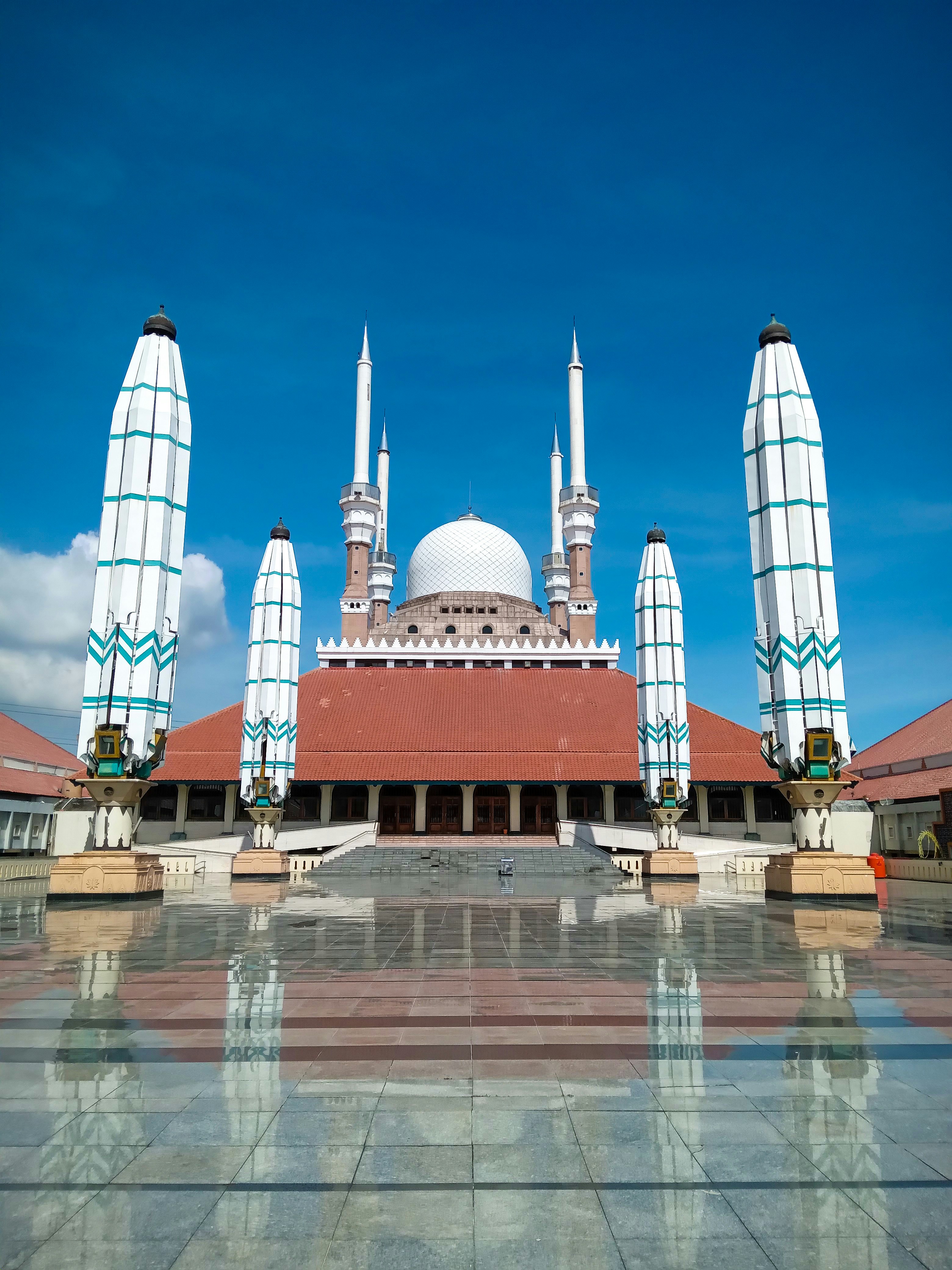 Symmetrical mosque complex featuring a white central dome, slender striped minarets, and a reflective marble plaza under a vivid blue sky.