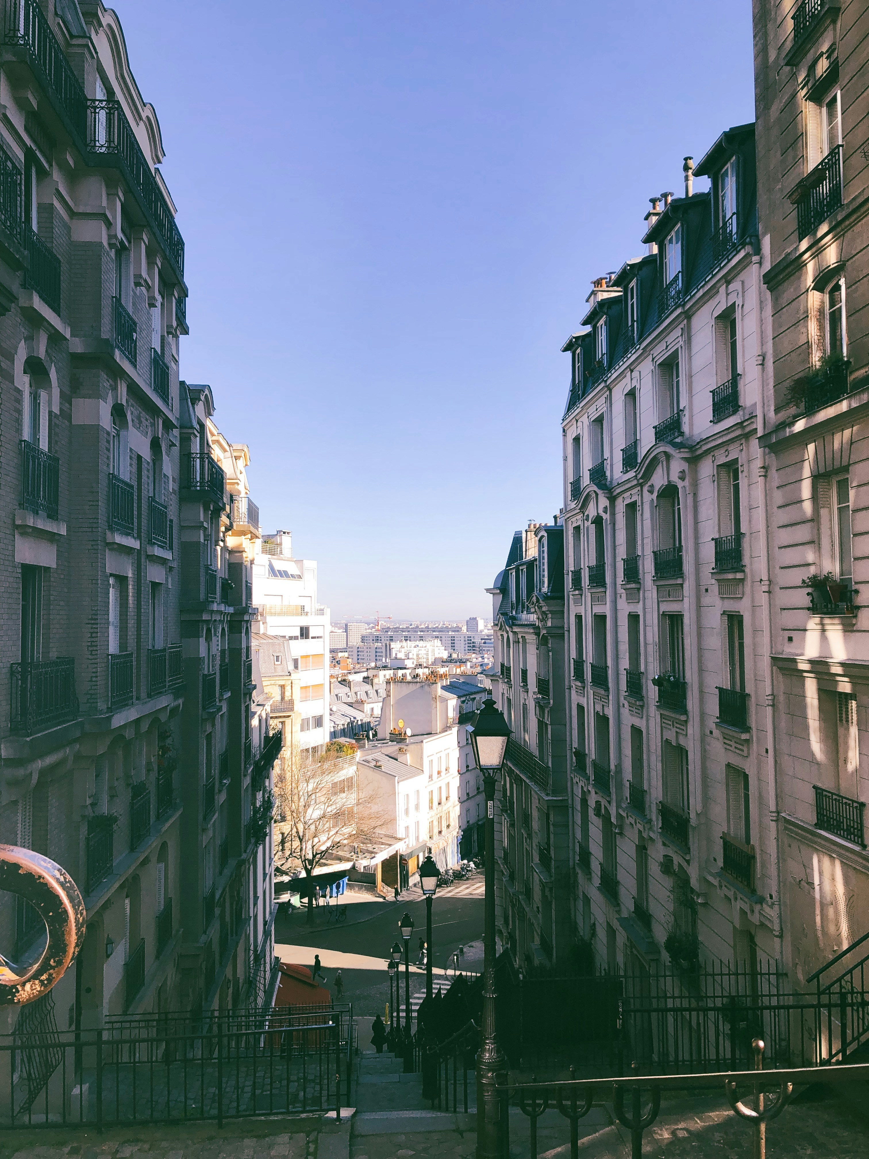 Narrow street in Montmartre framed by classic Parisian architecture, leading to a distant cityscape under a clear blue sky.
