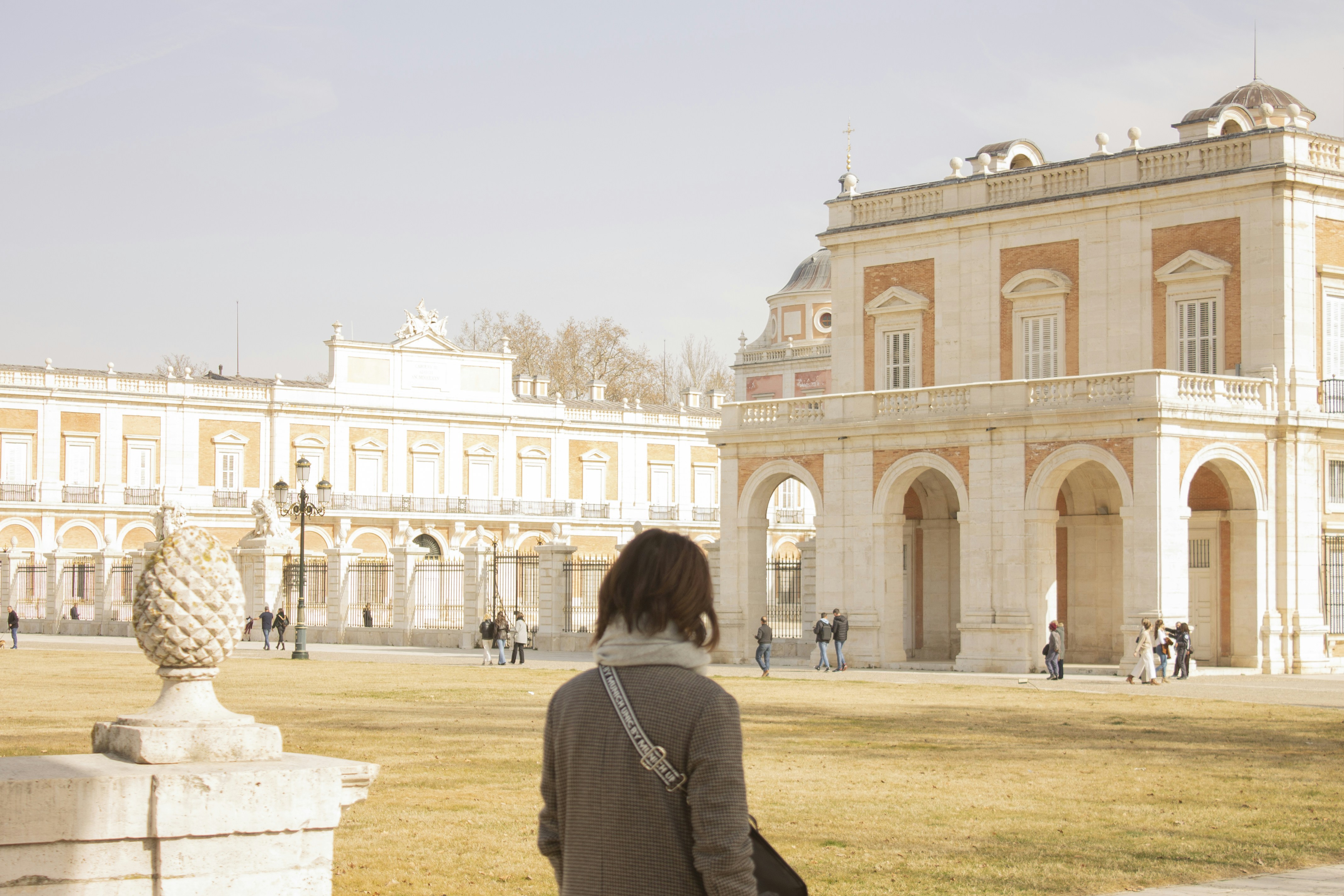 A woman gazes at the grand architecture of a historic building, with soft sunlight illuminating the scene. People stroll in the background, adding life to the serene atmosphere.