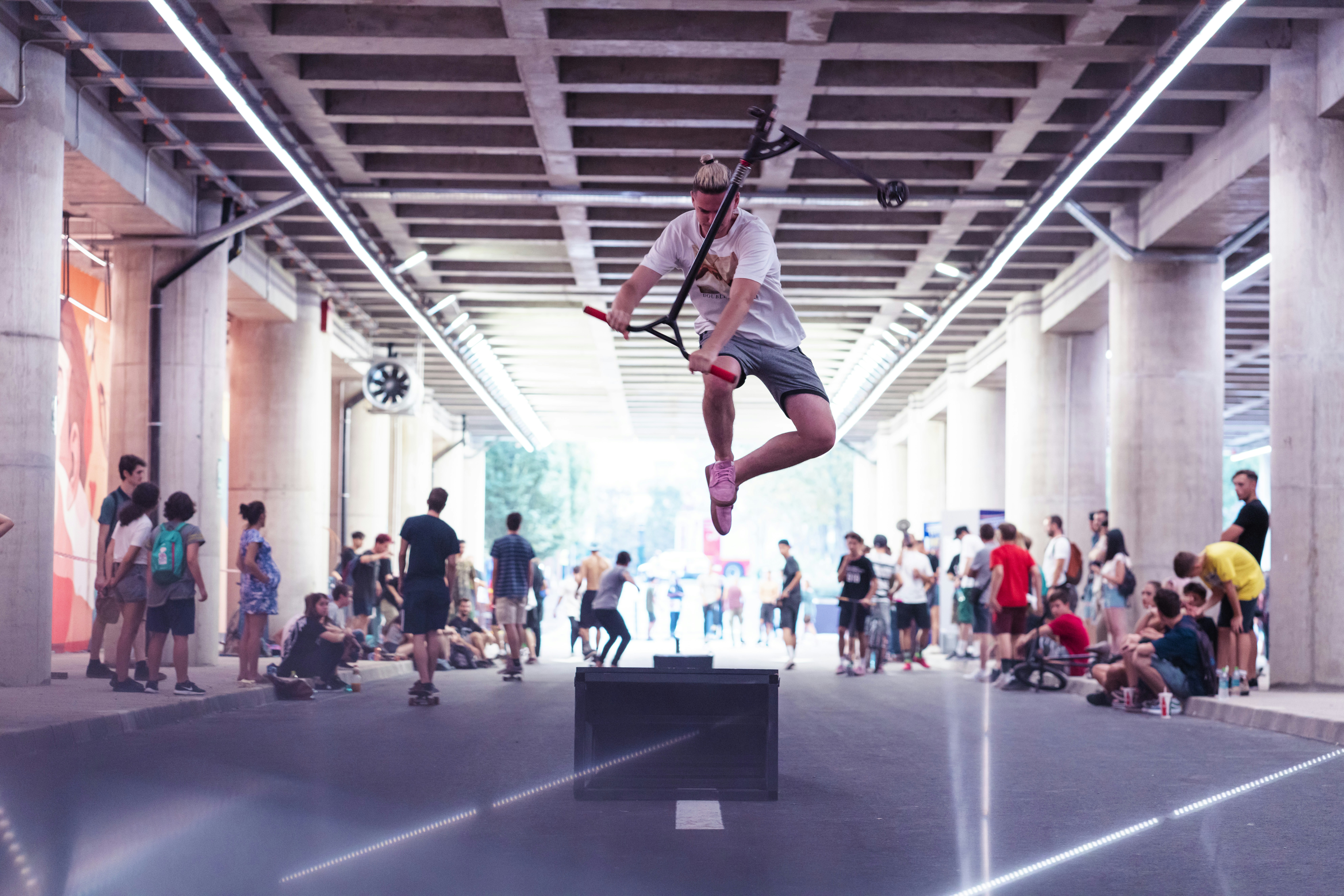 Street performer executing a mid-air trick on a scooter under an urban overpass, surrounded by an attentive crowd.