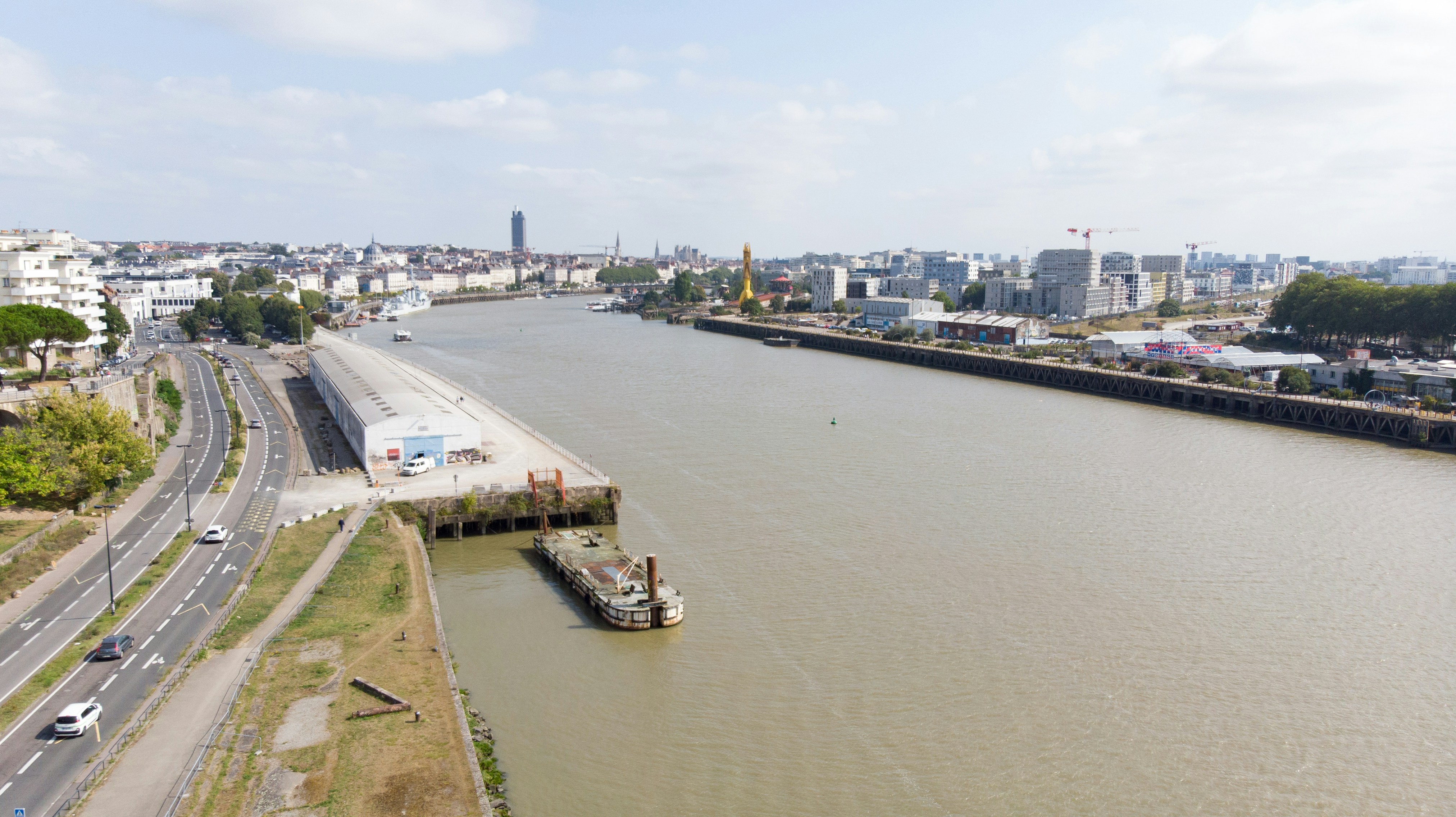 Wide river flowing through a cityscape with roads and industrial buildings under a cloudy sky.