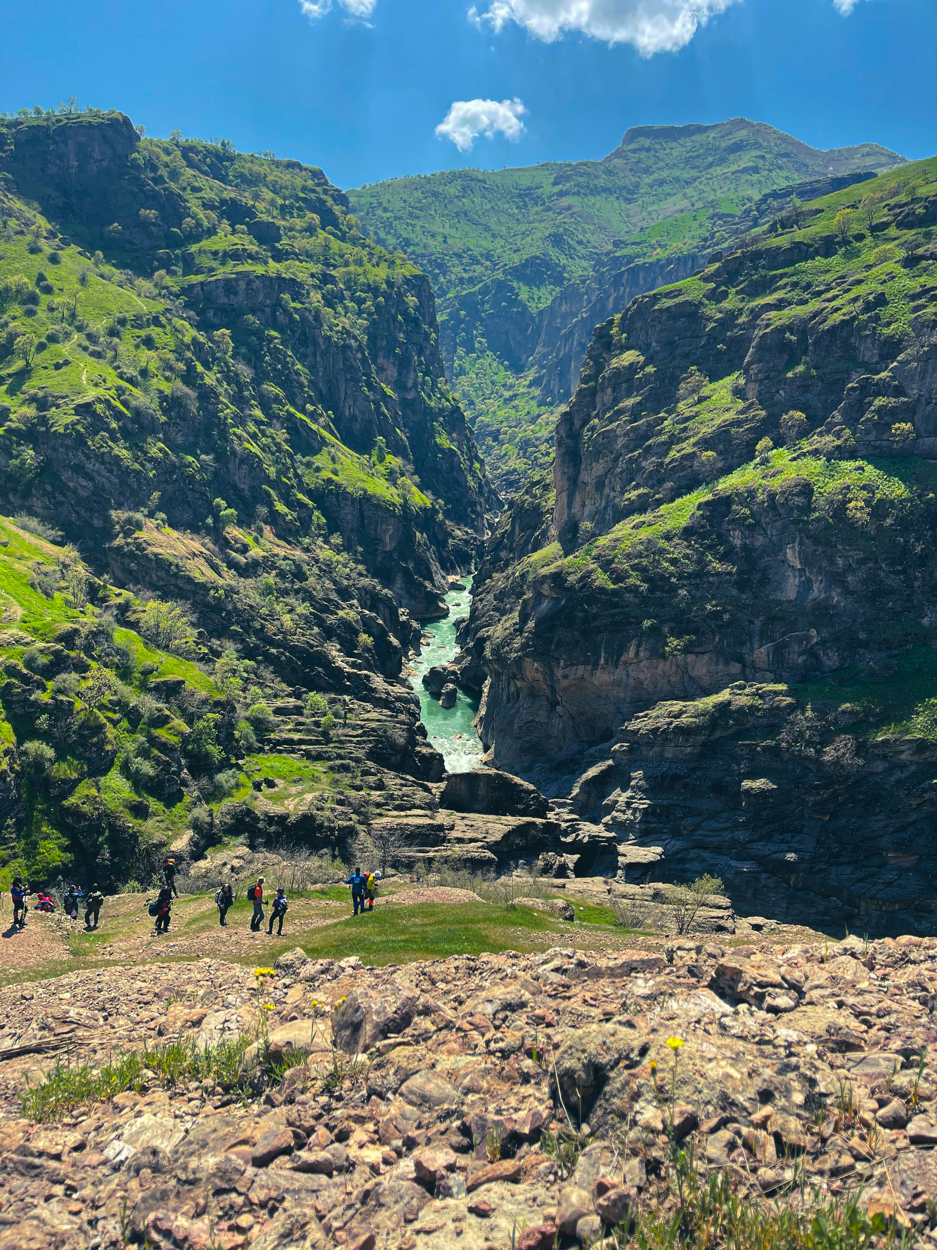A vibrant landscape showcasing a winding river flanked by steep cliffs, with hikers exploring the lush greenery. The scene captures the essence of adventure in a rugged terrain.