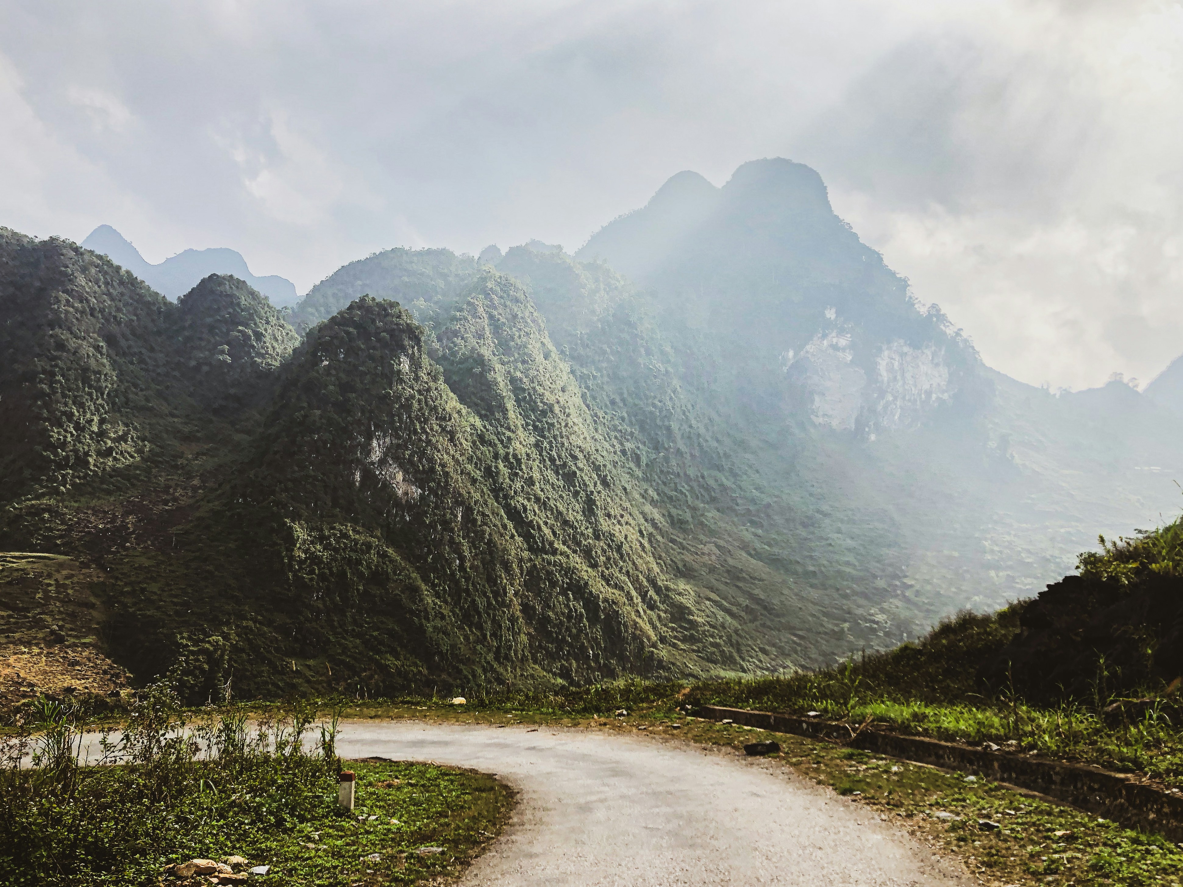 Curving road winds through lush green mountains under a cloudy sky, with sunlight streaming through the mist. 