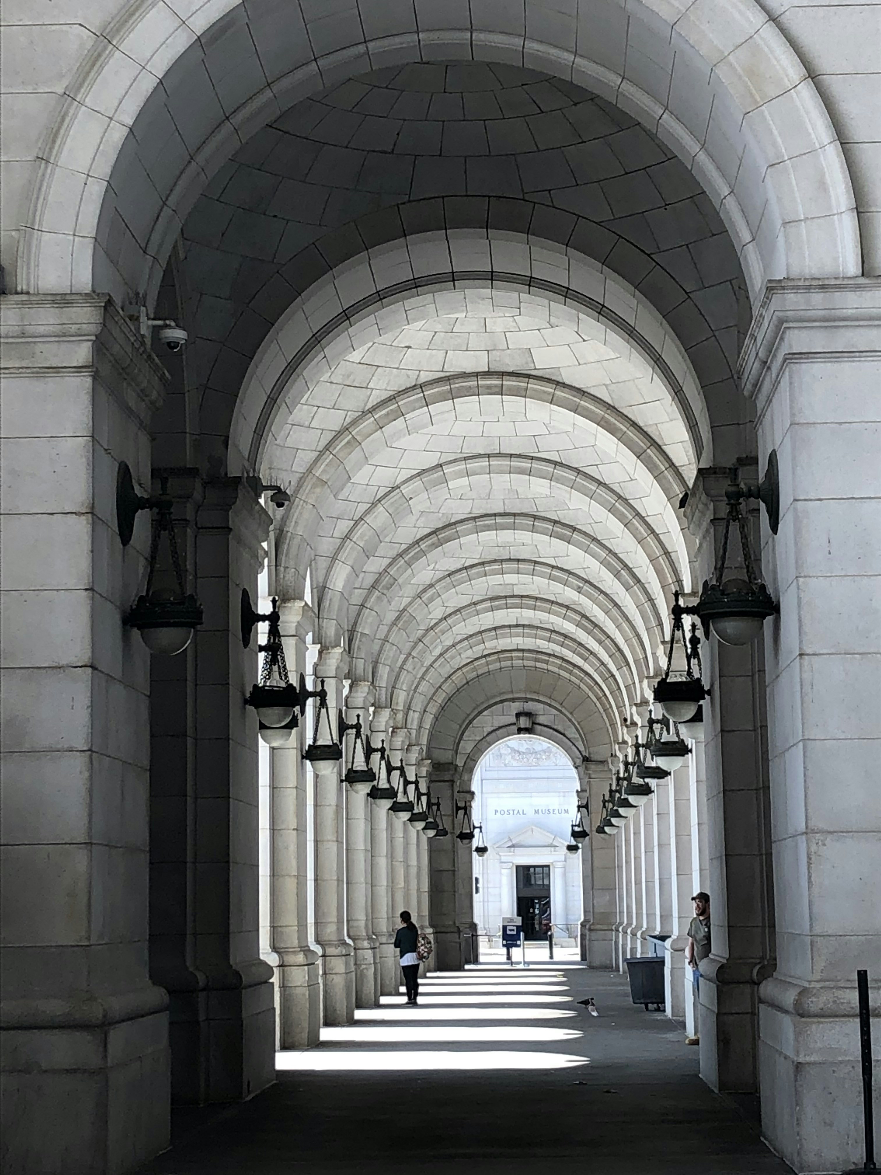 Architectural arches create a rhythmic pattern, leading to a distant entrance with figures walking through the corridor.