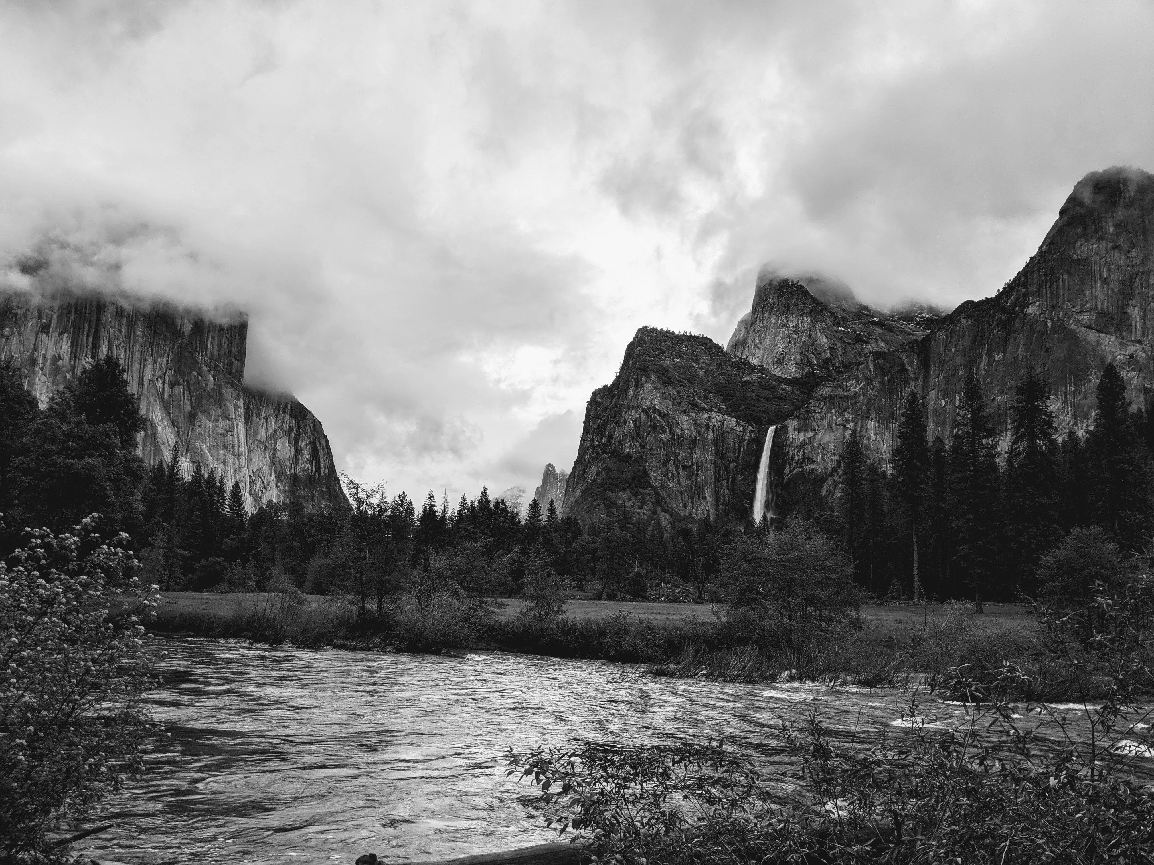 Dramatic black and white landscape showcasing towering cliffs and a waterfall shrouded in mist, framed by dense forest and a winding river.