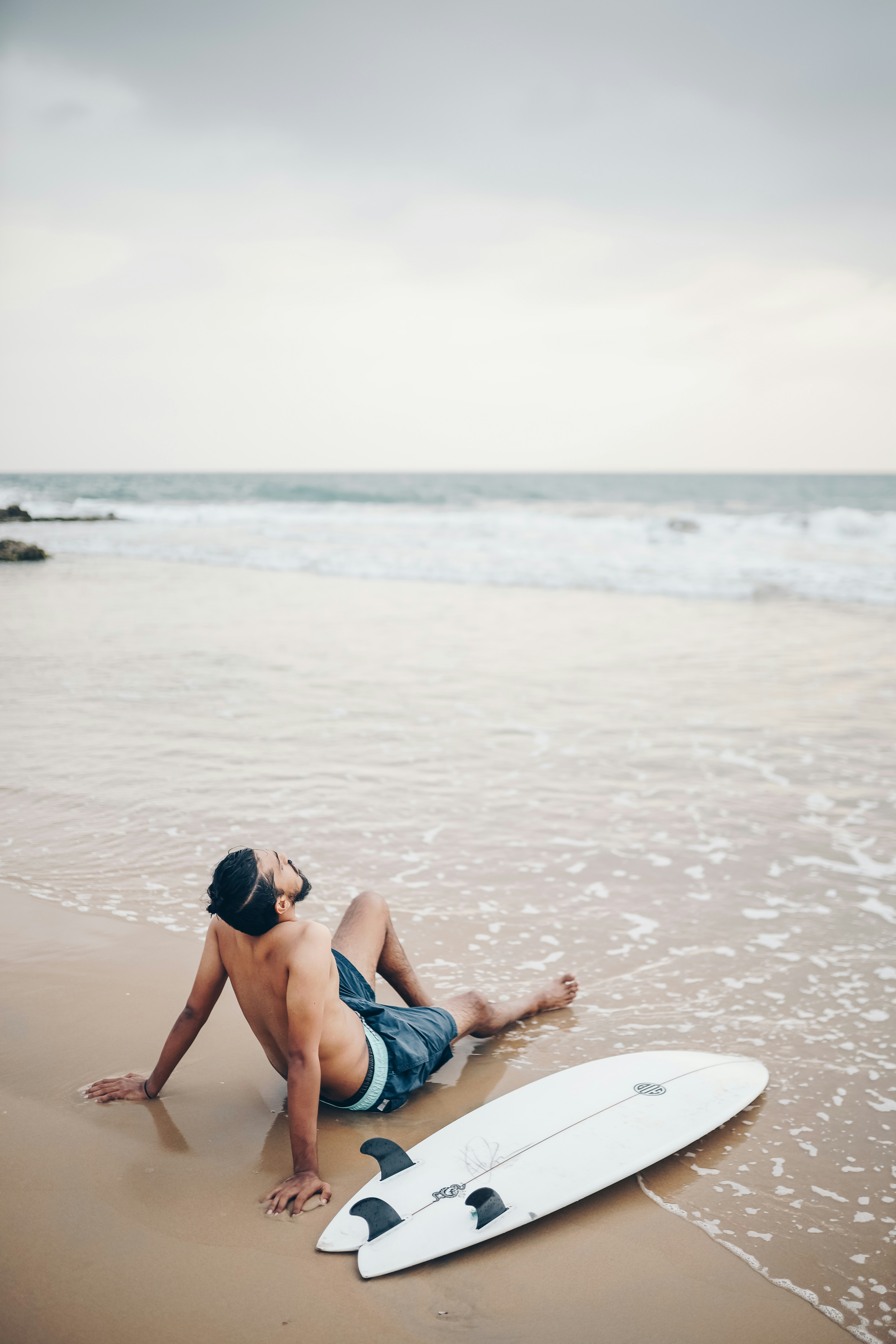 A surfer relaxing on the beach, gazing at the horizon with a surfboard beside him. The gentle waves create a serene atmosphere.