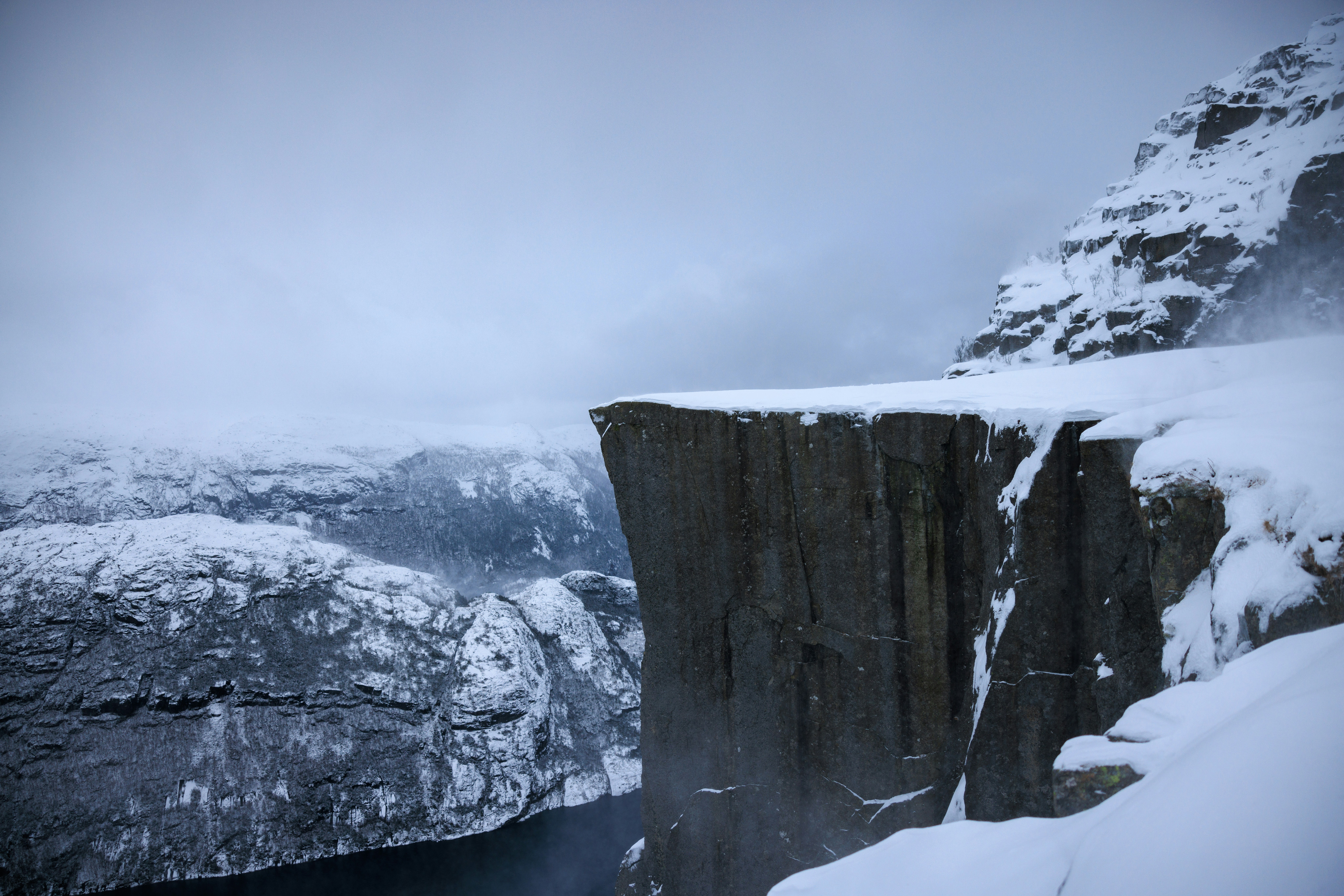 Snow-covered cliff edge overlooking a serene, icy fjord under a moody sky.