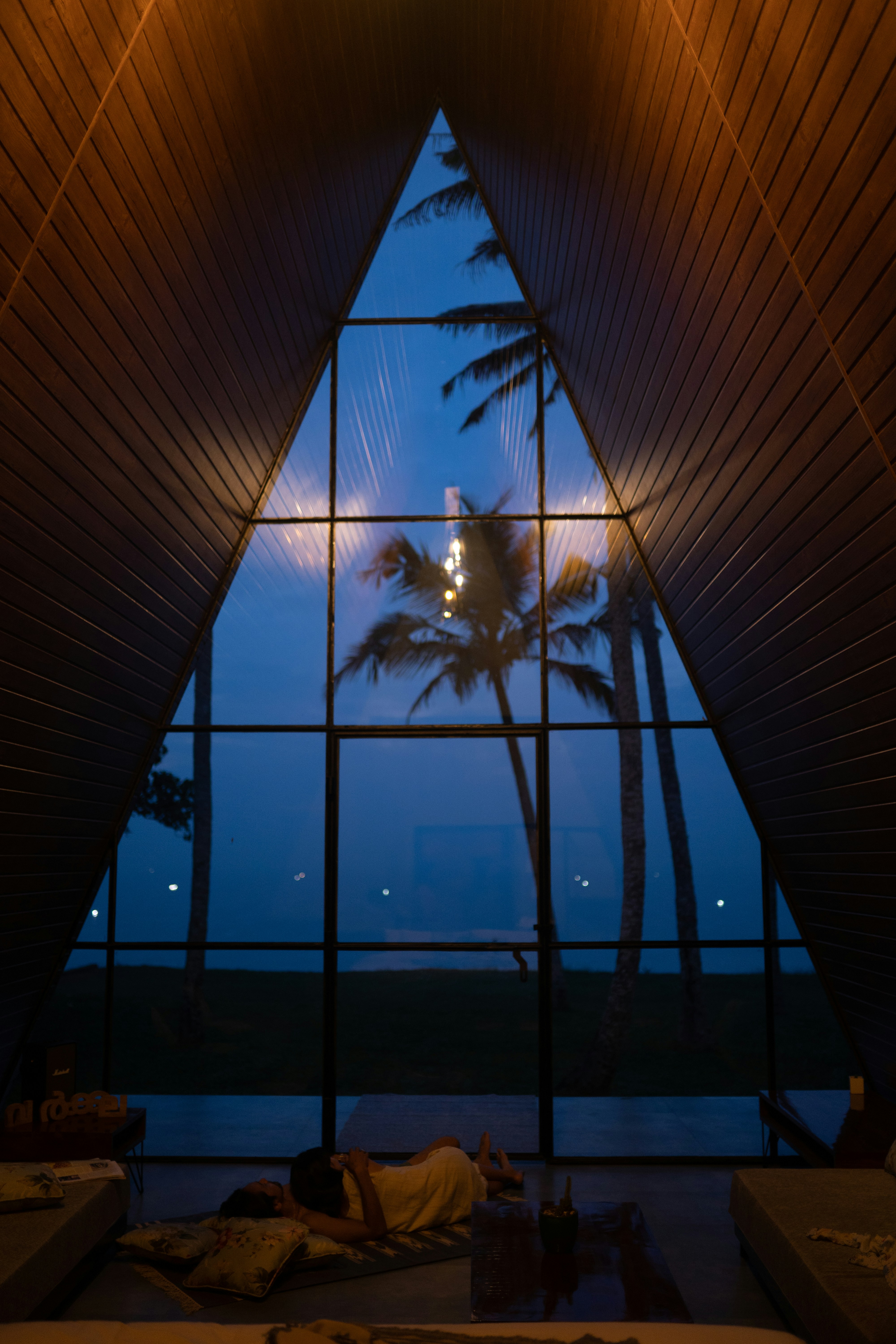 A serene interior space with A-frame architecture, featuring large glass windows that frame palm trees against a twilight sky. Soft lighting adds warmth to the cozy atmosphere.