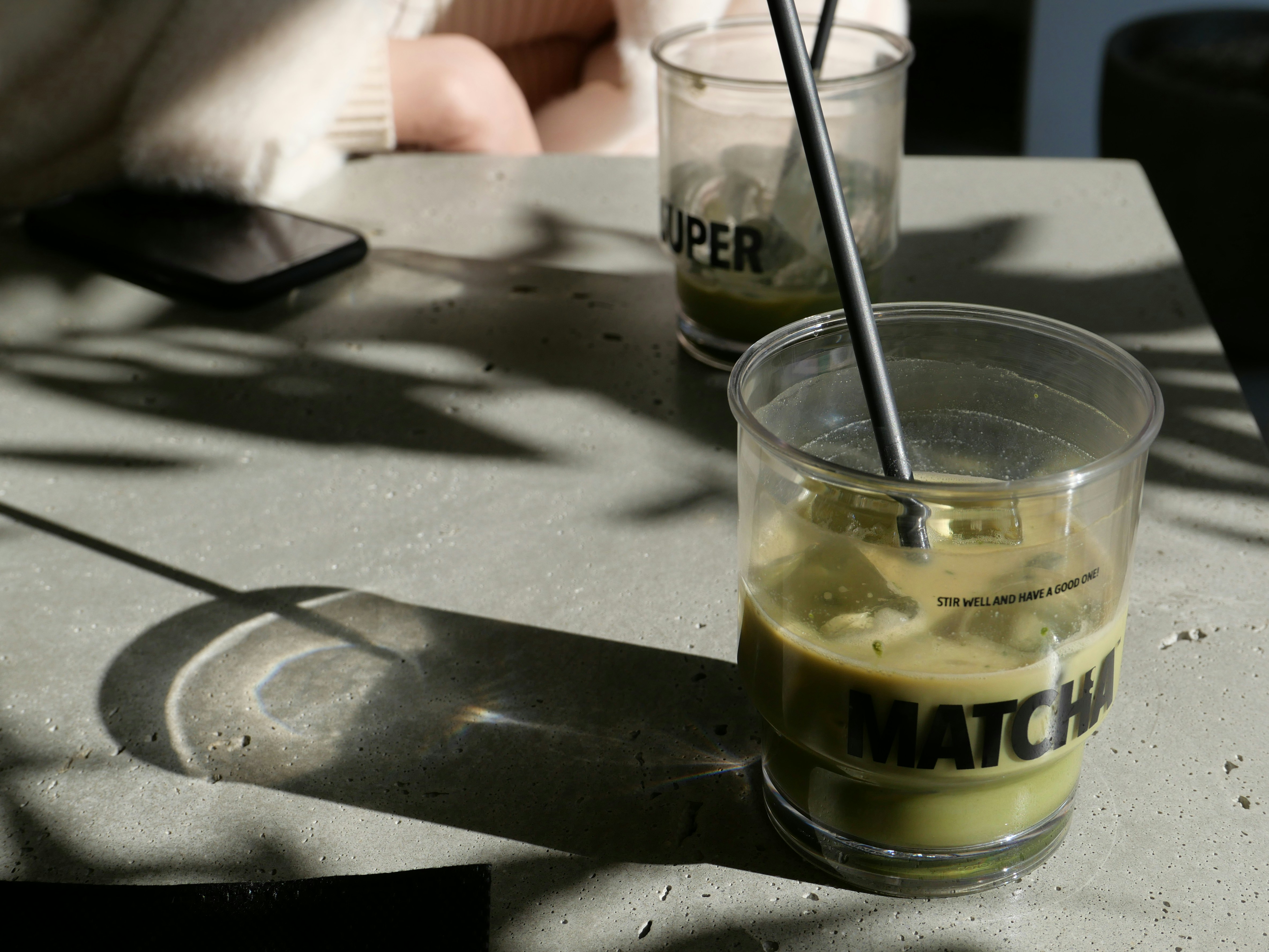 Two glasses of iced matcha on a textured table, with soft shadows and a hint of casual elegance. Sunlight filters through, enhancing the tranquil atmosphere.