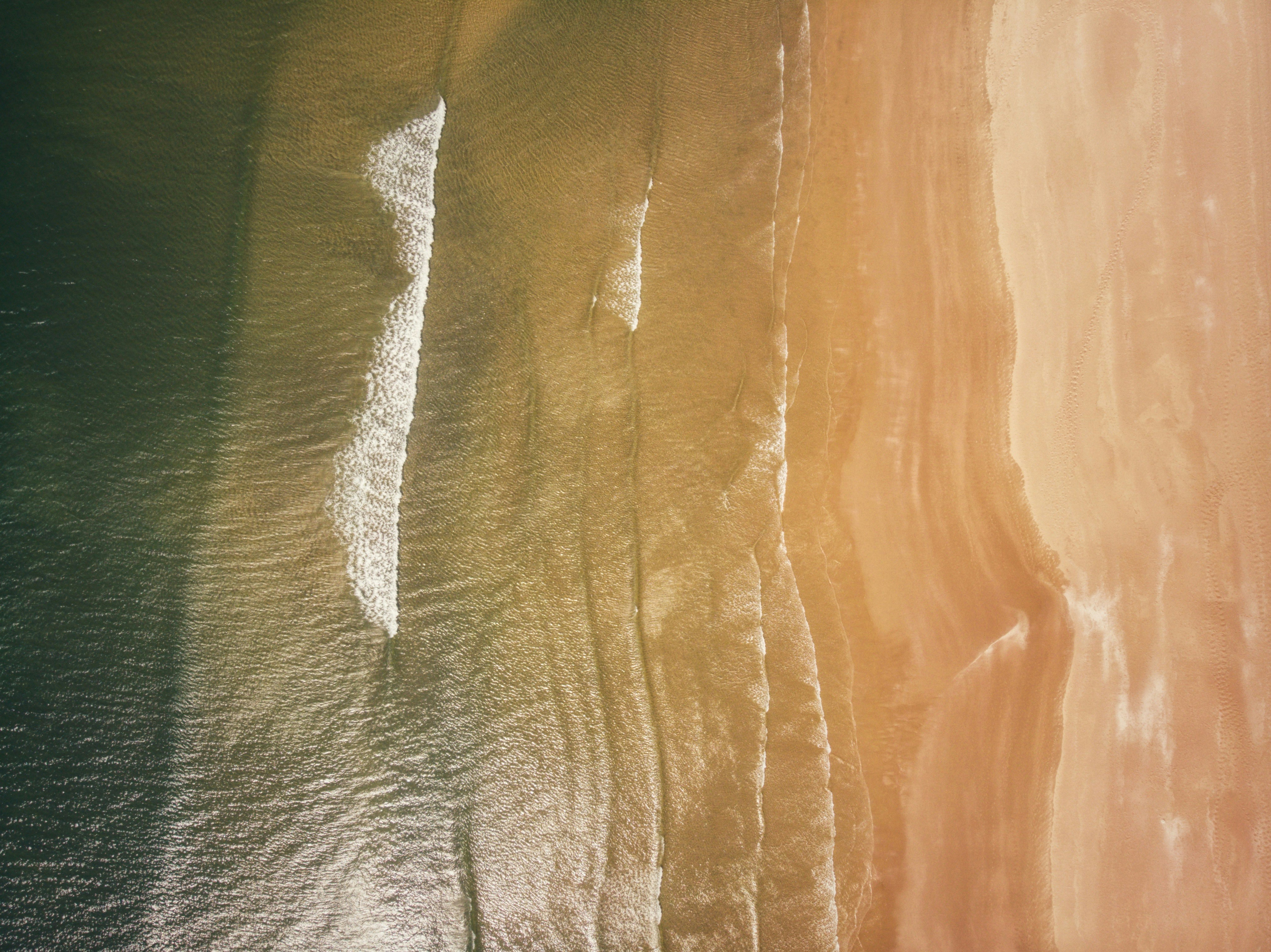 Aerial view of waves gently lapping against a sandy beach, showcasing the gradient of colors from deep blue to golden sand. The texture of the water and shore creates a harmonious visual rhythm.
