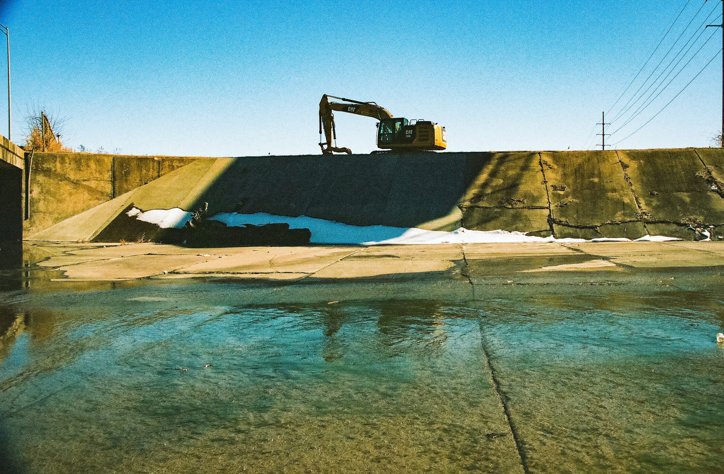 Excavator positioned on a concrete embankment with reflections in the water below, under a clear blue sky. The scene captures the juxtaposition of machinery and nature.