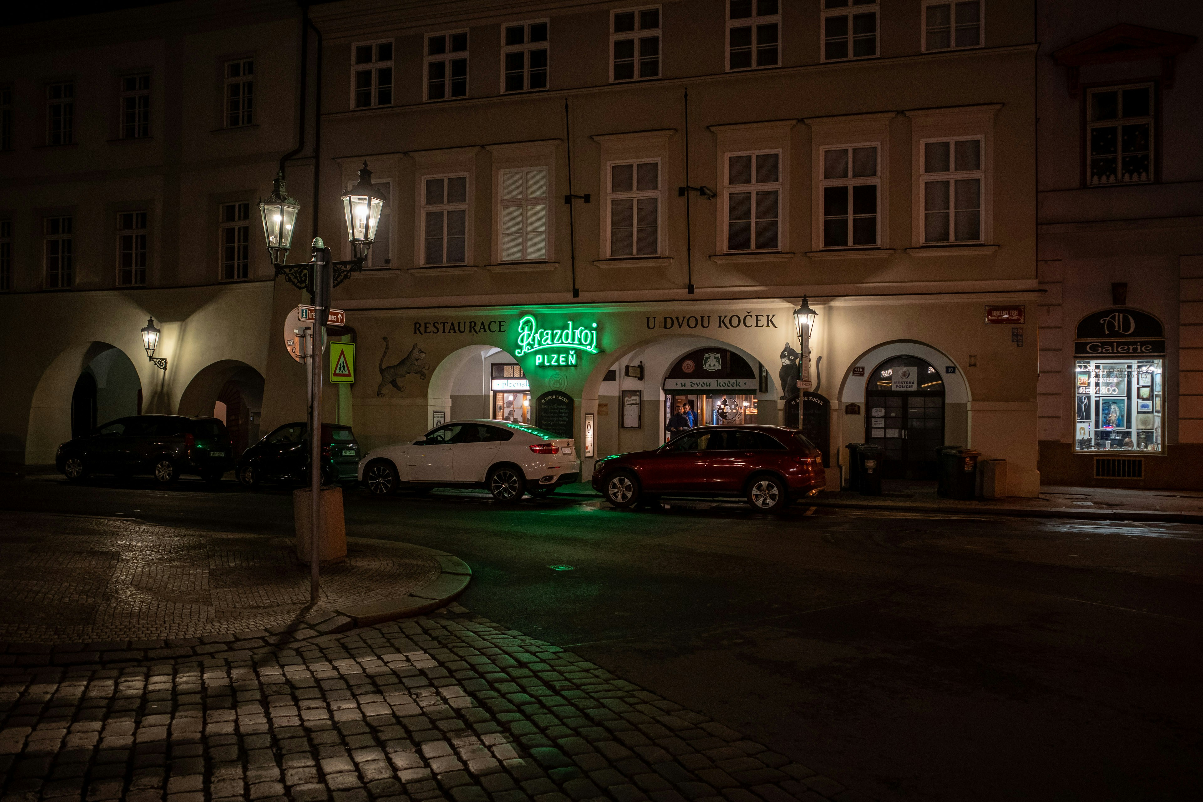 Cars parked in front of a historic building with glowing neon signage on a quiet cobblestone street at night.