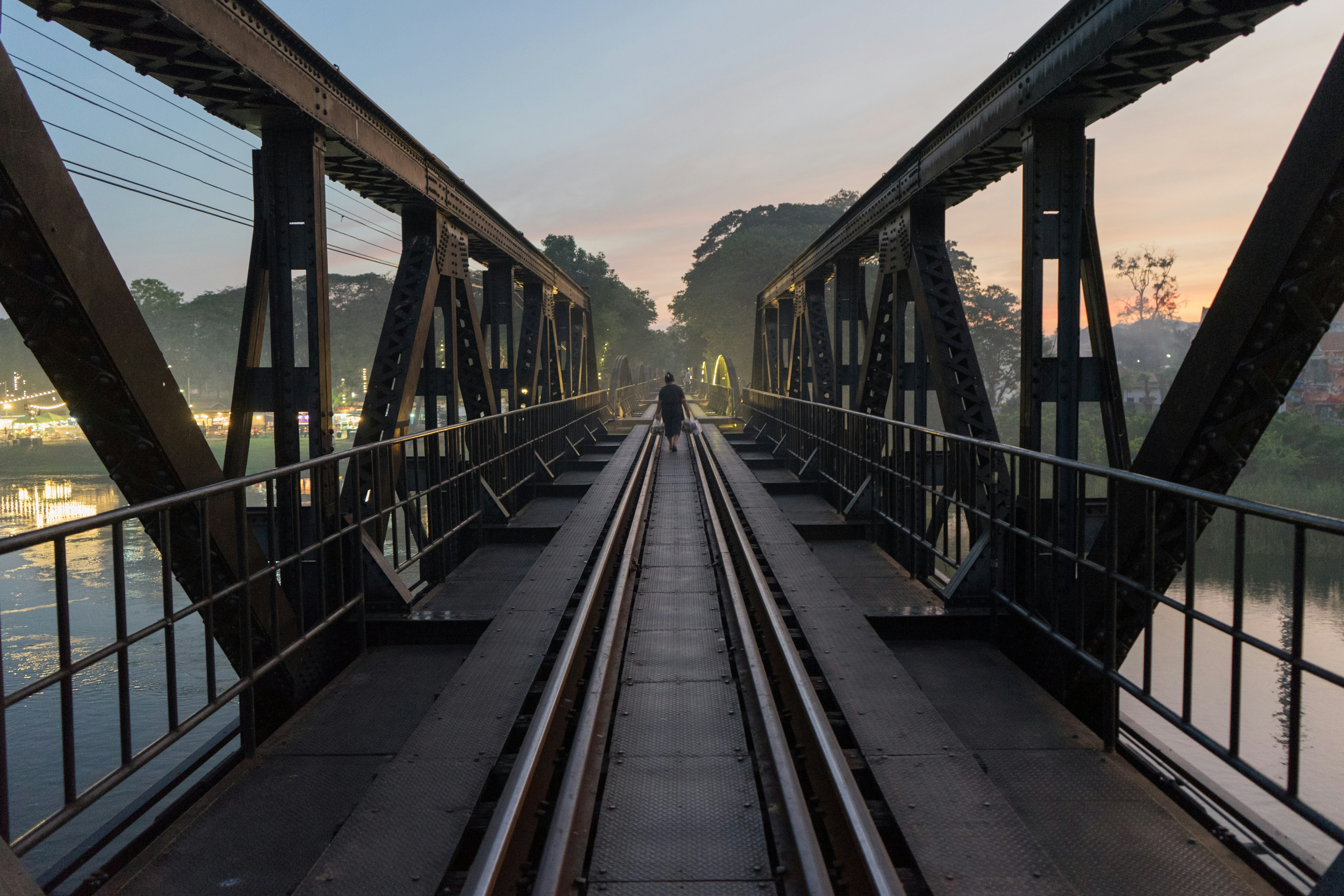 A lone figure walks along a railway bridge at dusk, framed by the industrial architecture and serene waters below.