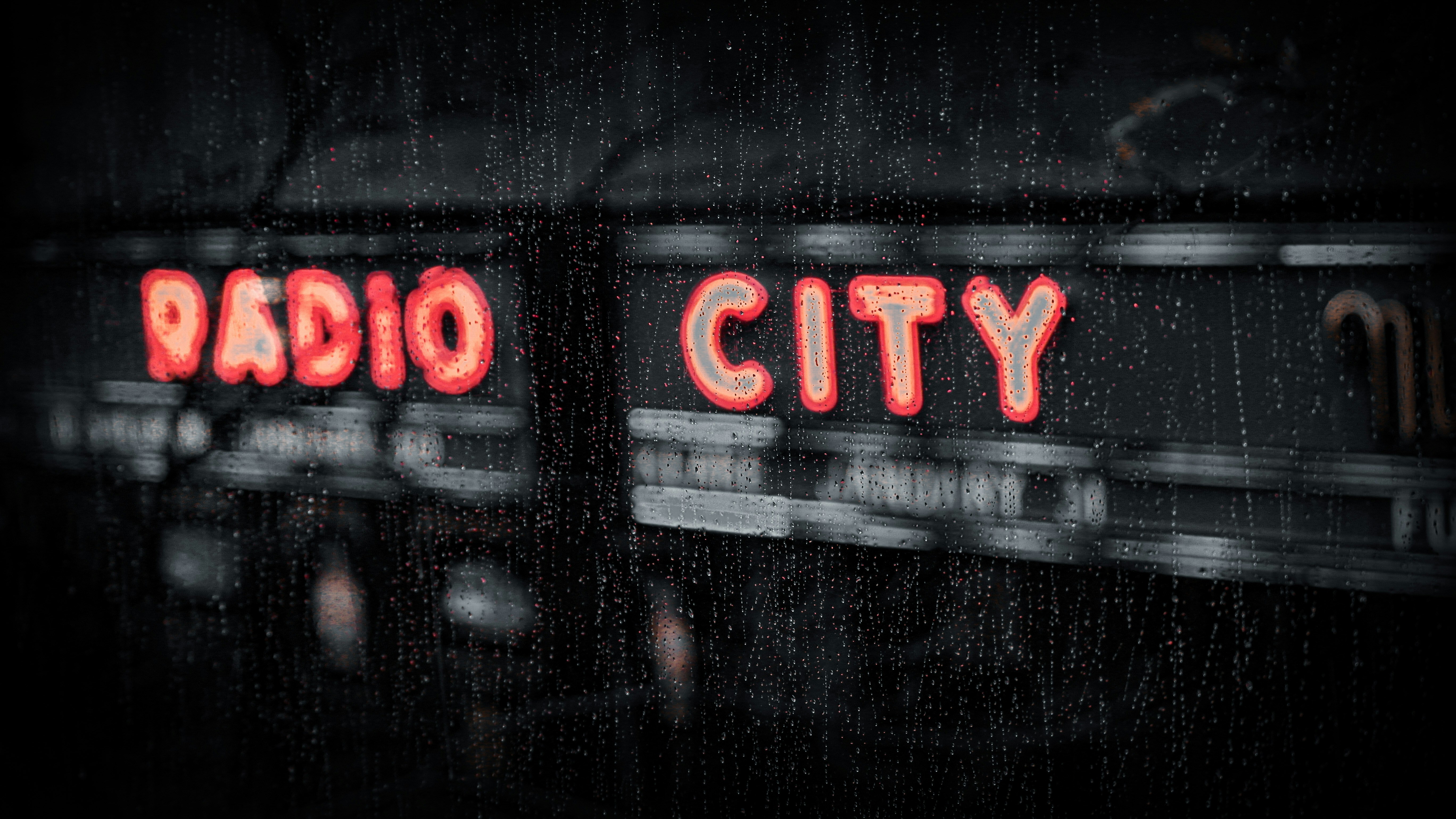 Red neon 'RADIO CITY' sign glows through rain on a dark storefront, with water droplets creating texture and bokeh in the foreground.