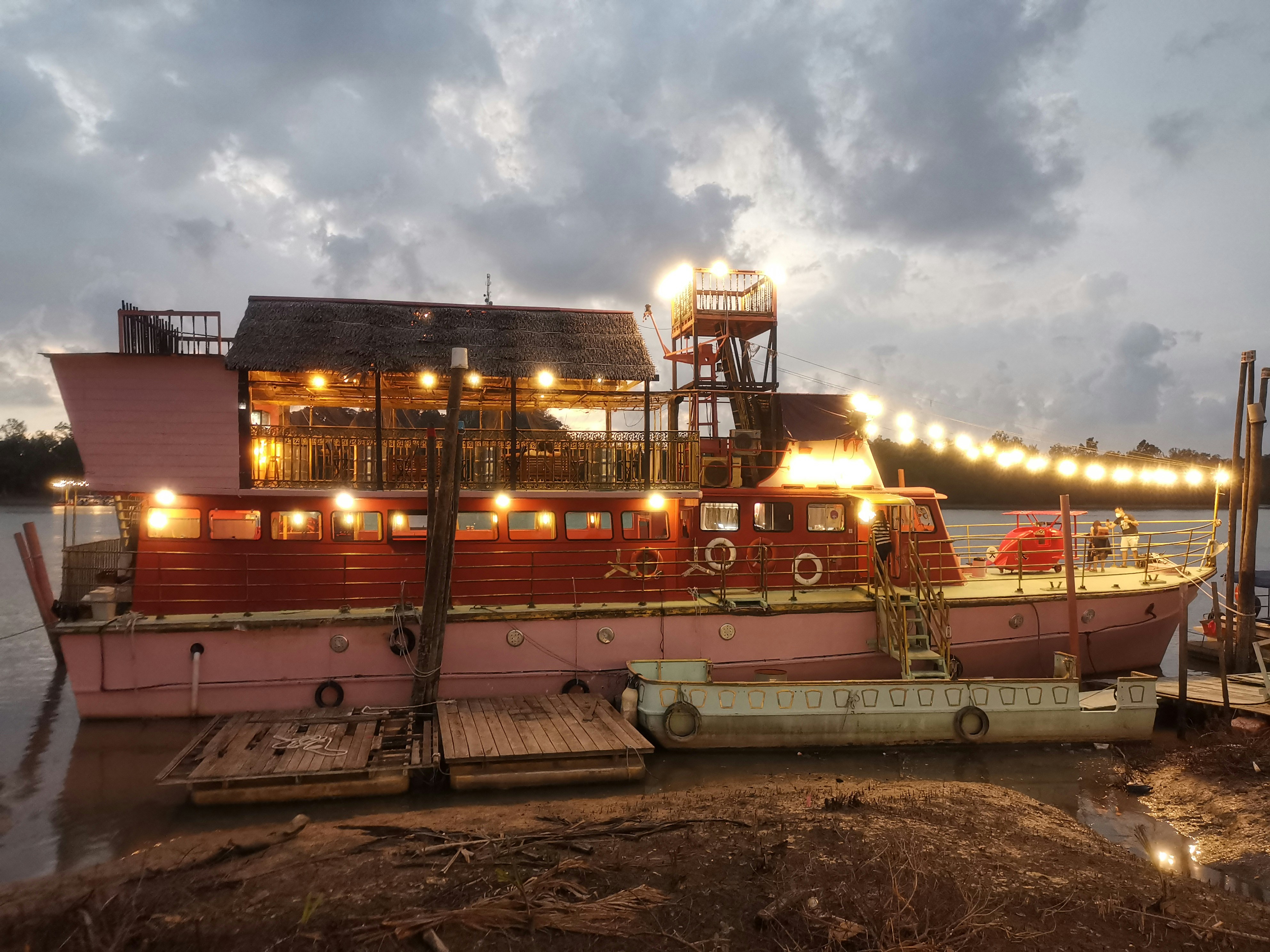 Illuminated boat docked by a riverbank under a cloudy evening sky.
