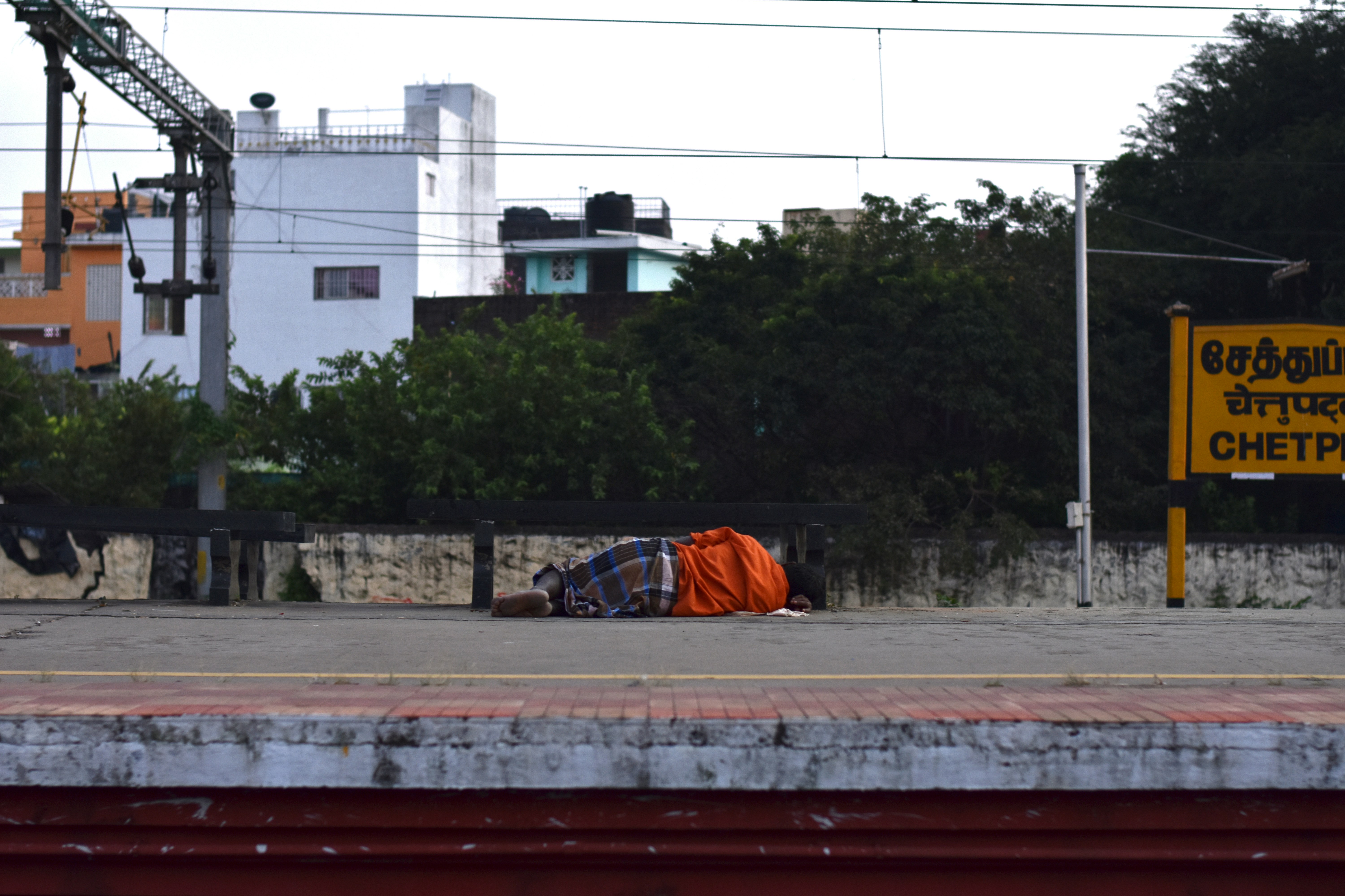 A person wrapped in an orange cloth lies on a train platform, surrounded by urban structures and greenery. The scene reflects the contrast between city life and moments of solitude.