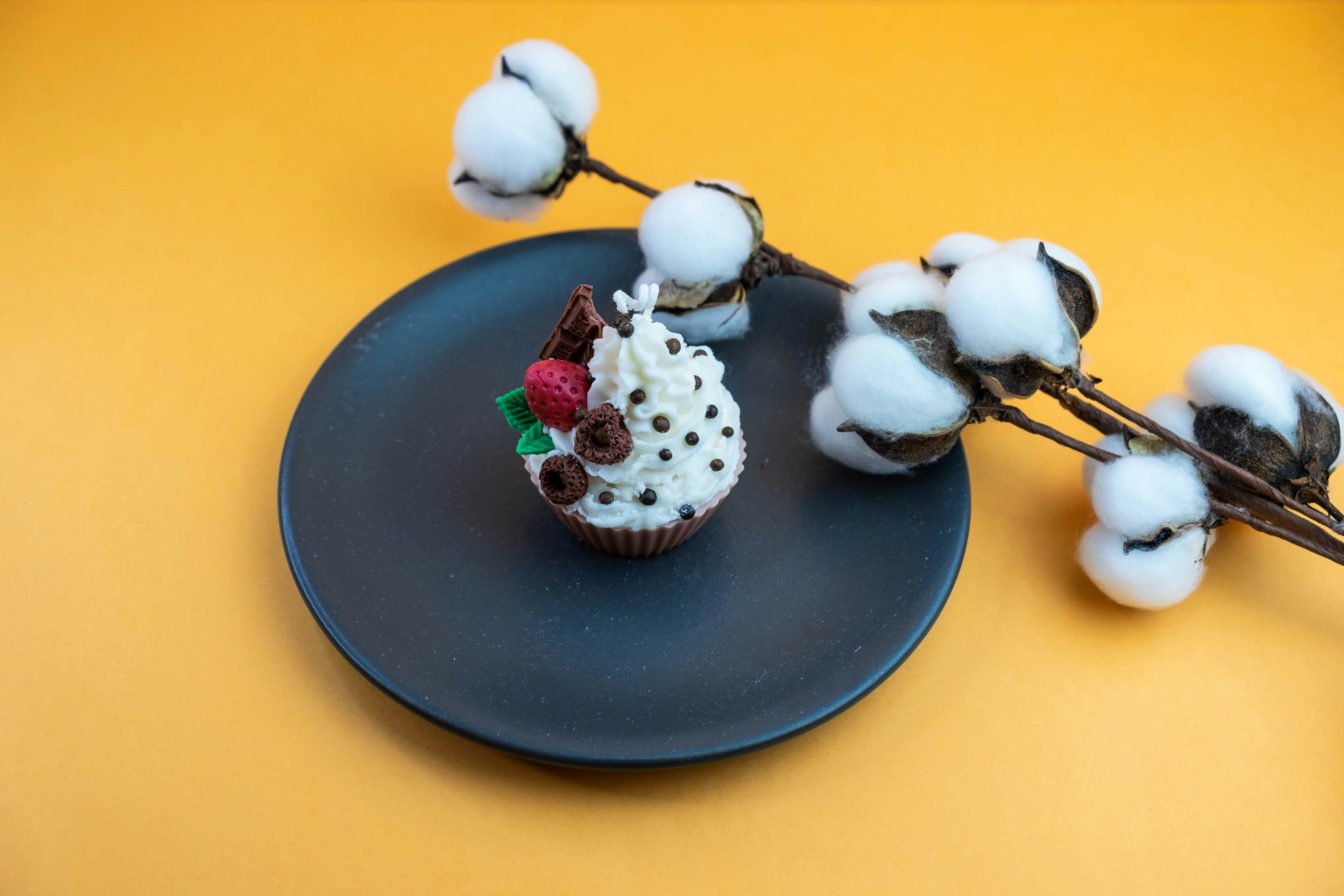 A whimsical cupcake topped with cream and decorative elements sits on a dark plate, accompanied by a branch of cotton. The vibrant background enhances the playful theme.