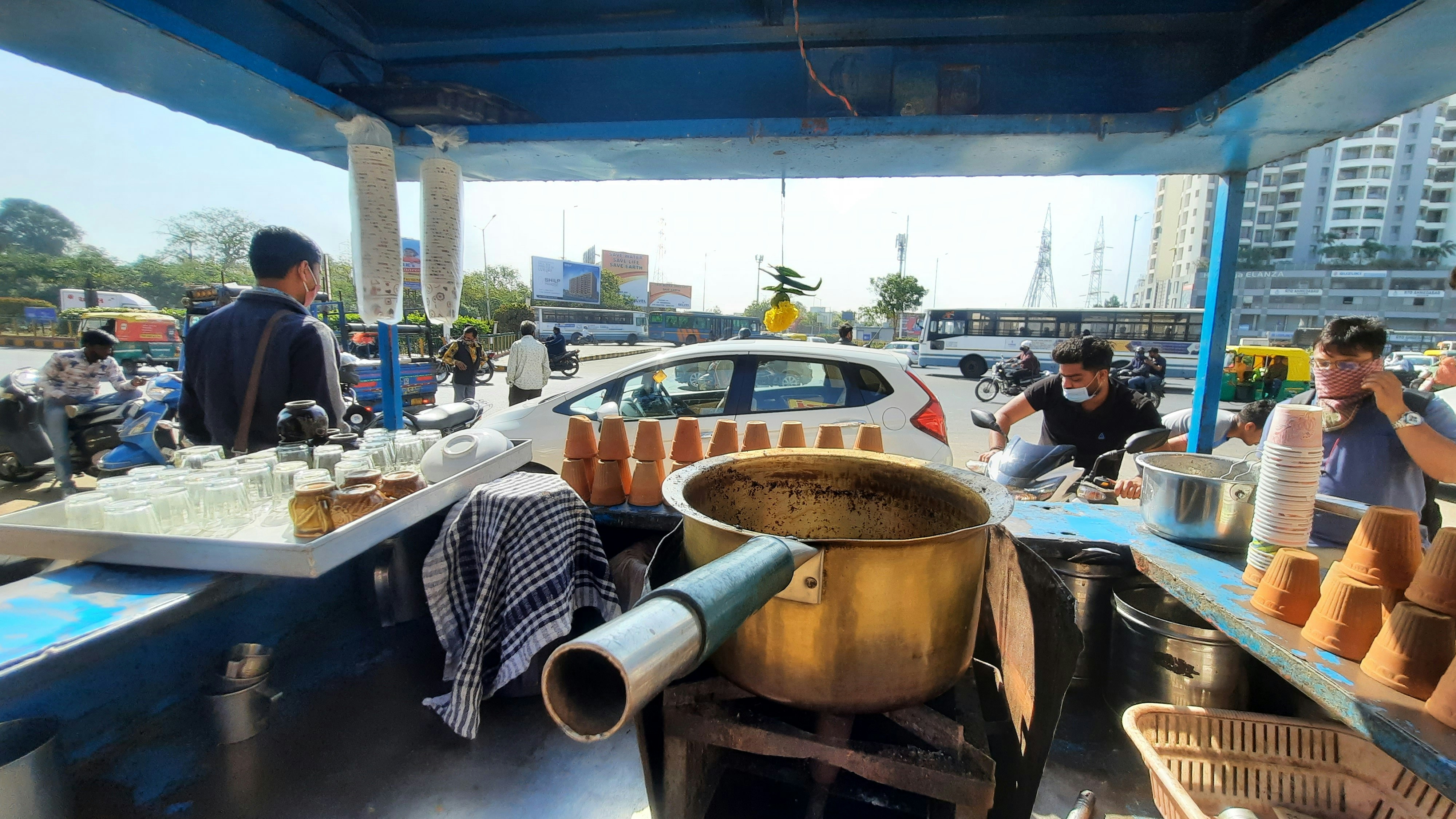 Busy street tea stall with patrons enjoying beverages and a chef preparing tea in a large pot.