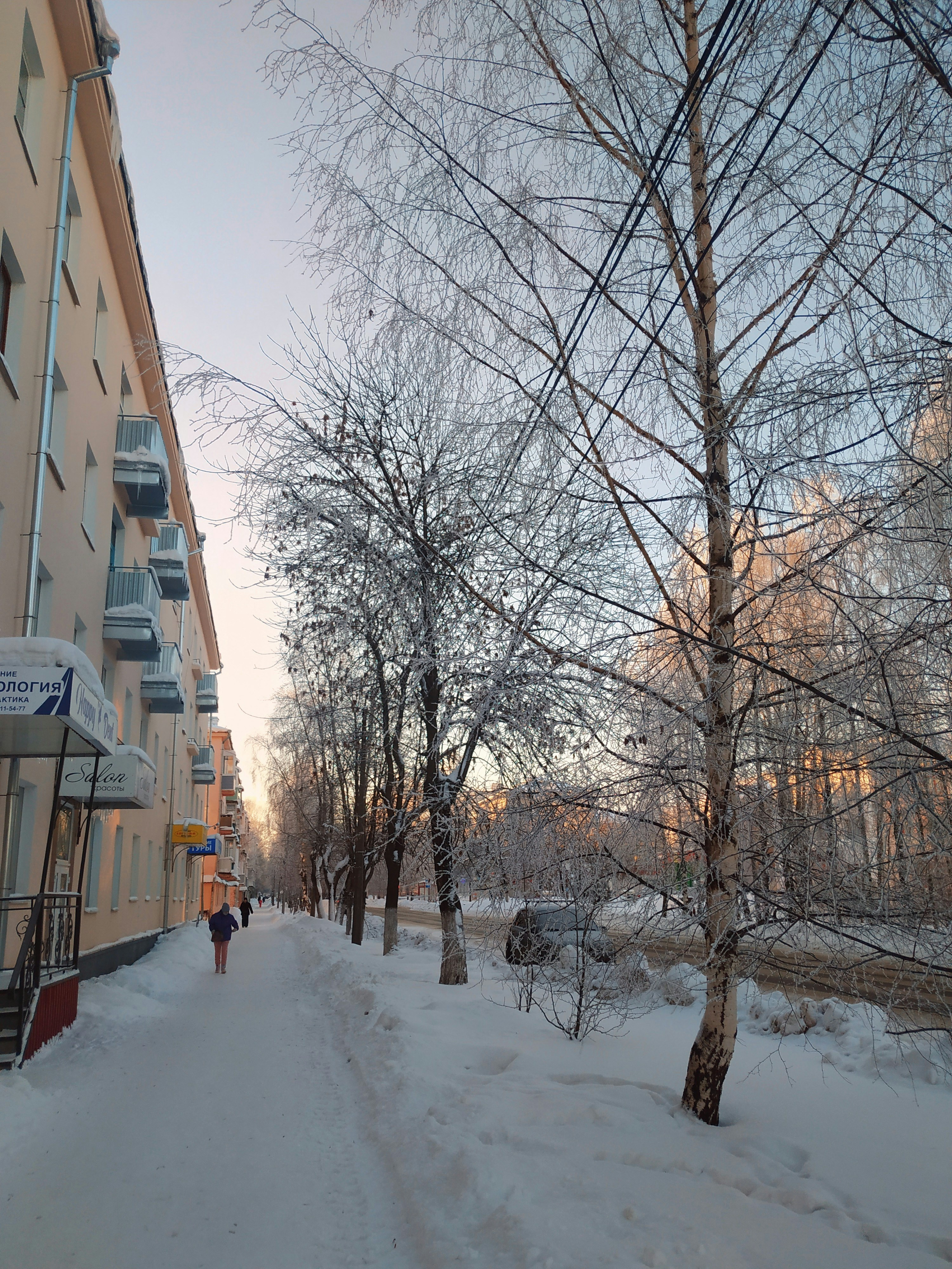 Snowy city sidewalk flanked by a pale yellow building and bare trees; distant pedestrians and a parked car along a sunlit street.