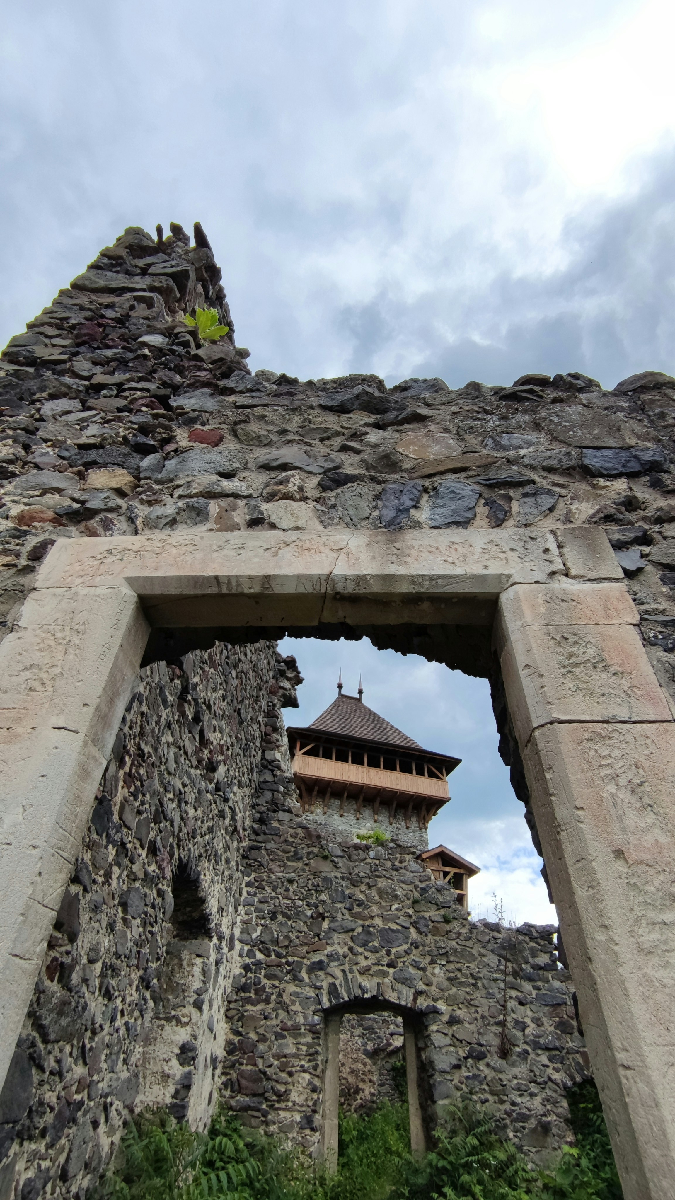 Ancient stone ruins with a partially visible wooden structure in the background, framed by an archway. Lush greenery peeks through the stones.