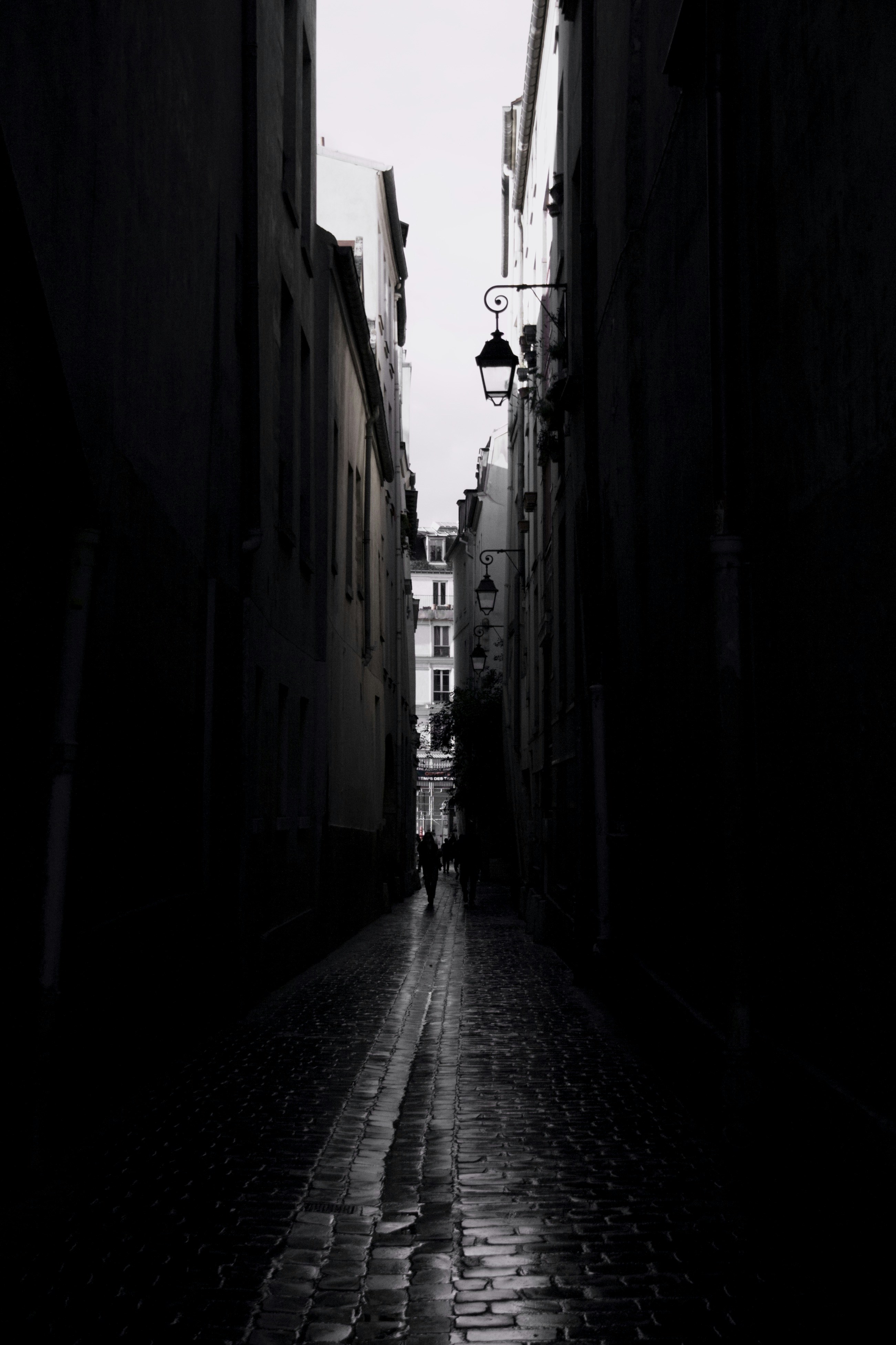 Dimly lit alleyway with cobblestone pavement reflecting light, flanked by tall buildings. A solitary figure walks towards the distant light.