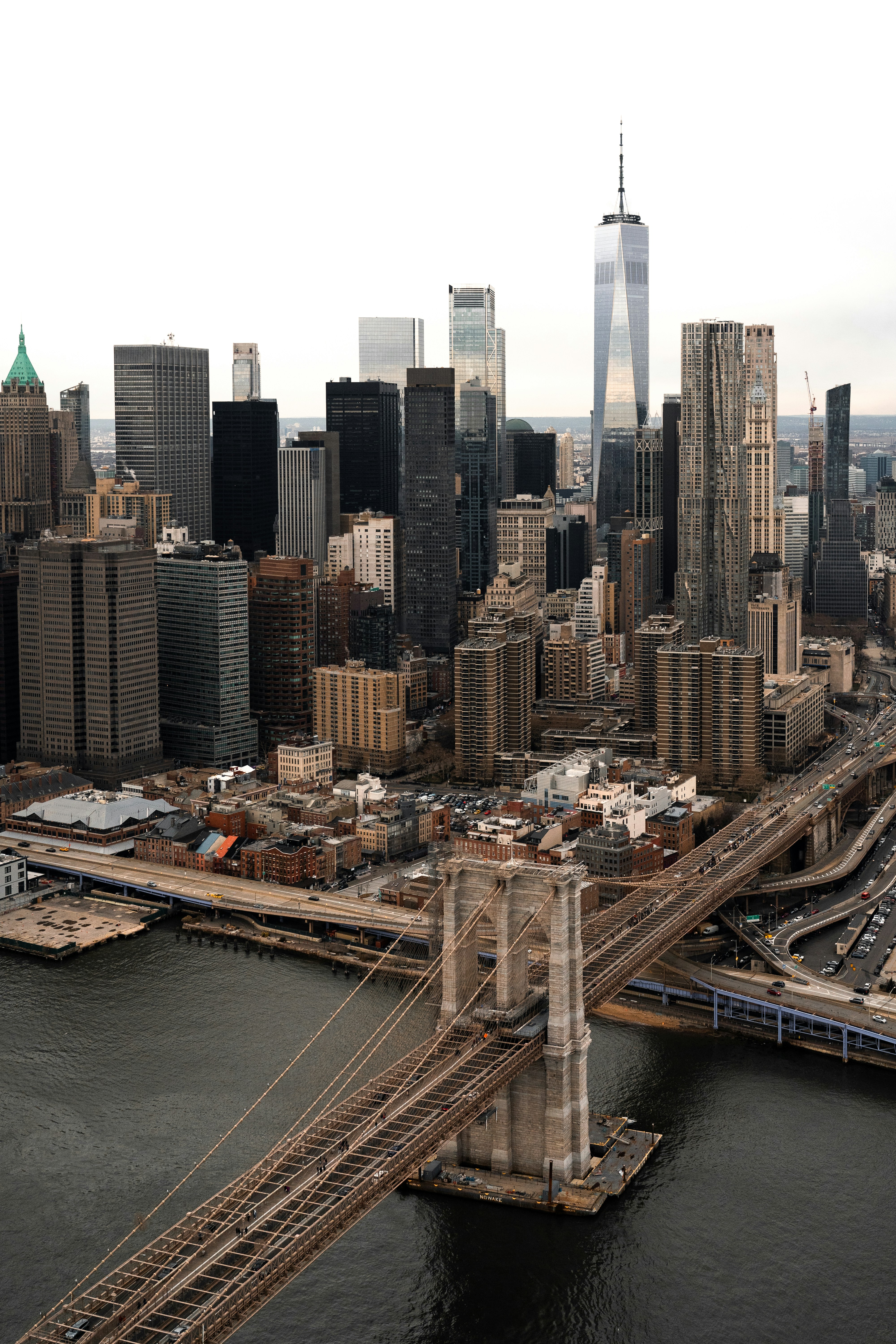 Aerial view of the Brooklyn Bridge connecting Manhattan and Brooklyn, with skyscrapers and the One World Trade Center in the background.