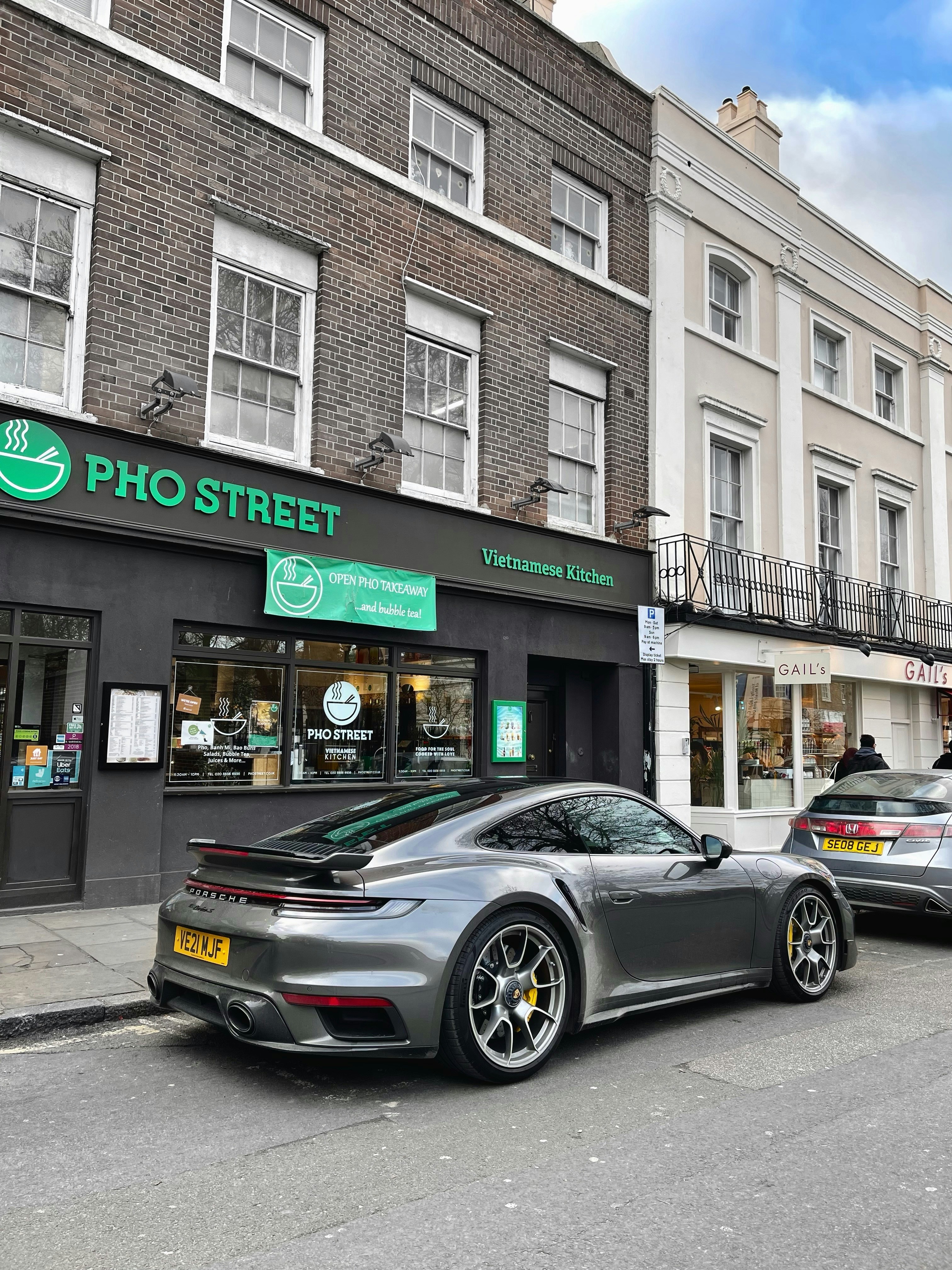 A sleek gray Porsche 911 parked beside a vibrant Vietnamese restaurant, showcasing the intersection of modern cuisine and luxury automobiles.