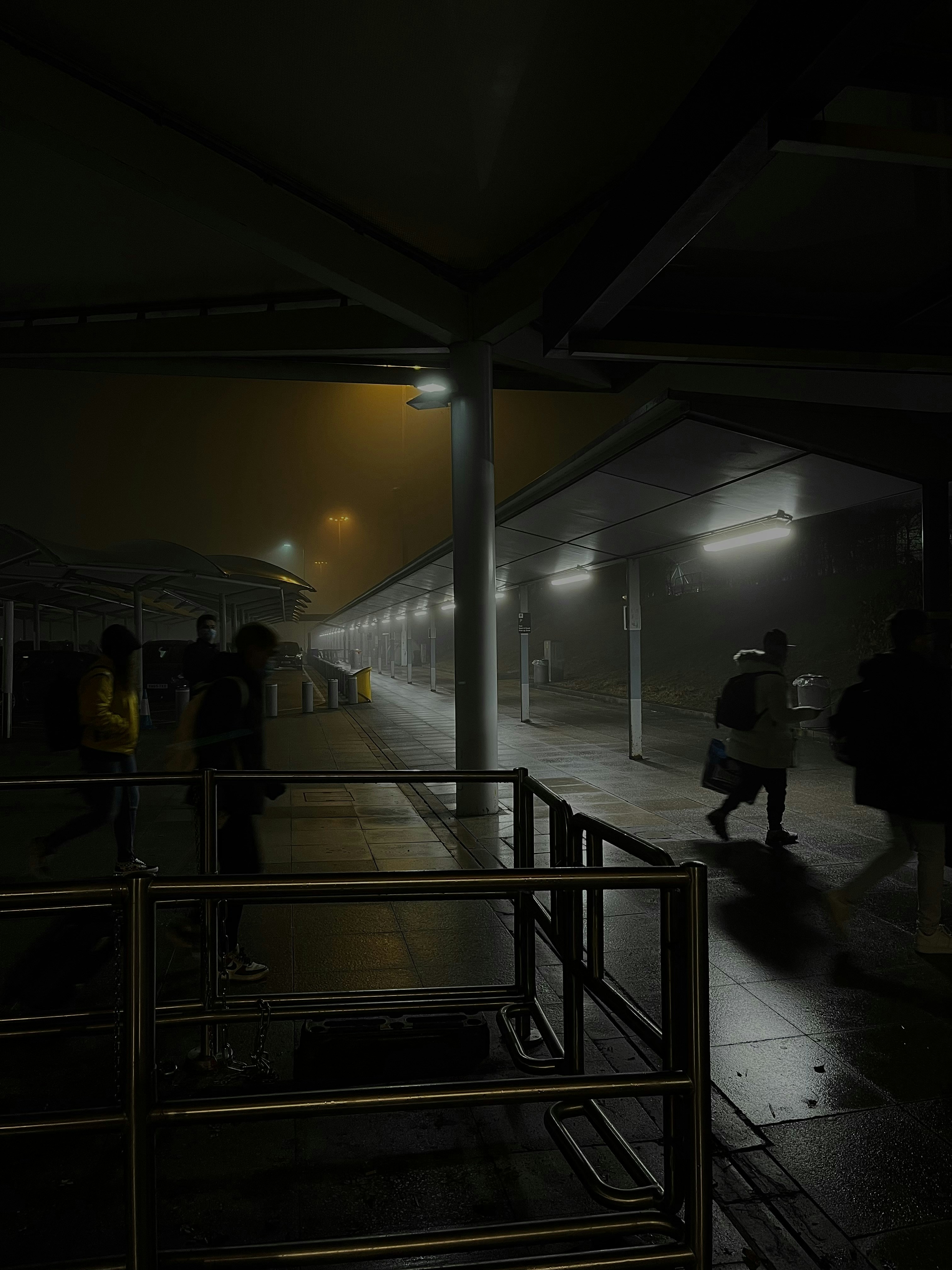 Dimly lit transit station enveloped in fog, with silhouettes of commuters moving through the misty atmosphere.