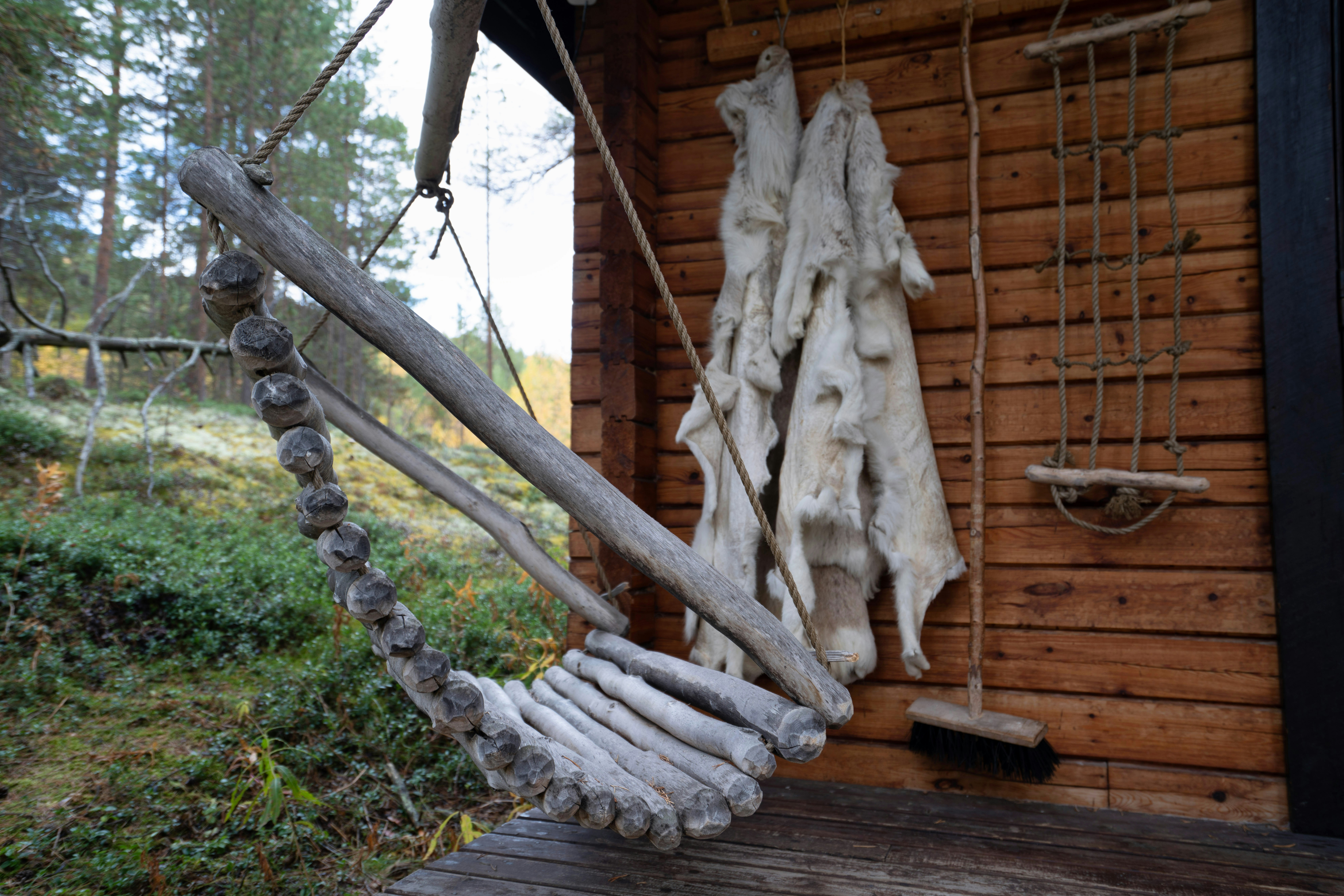 Wooden swing hanging from a cabin, surrounded by lush greenery and animal hides displayed on the wall. 