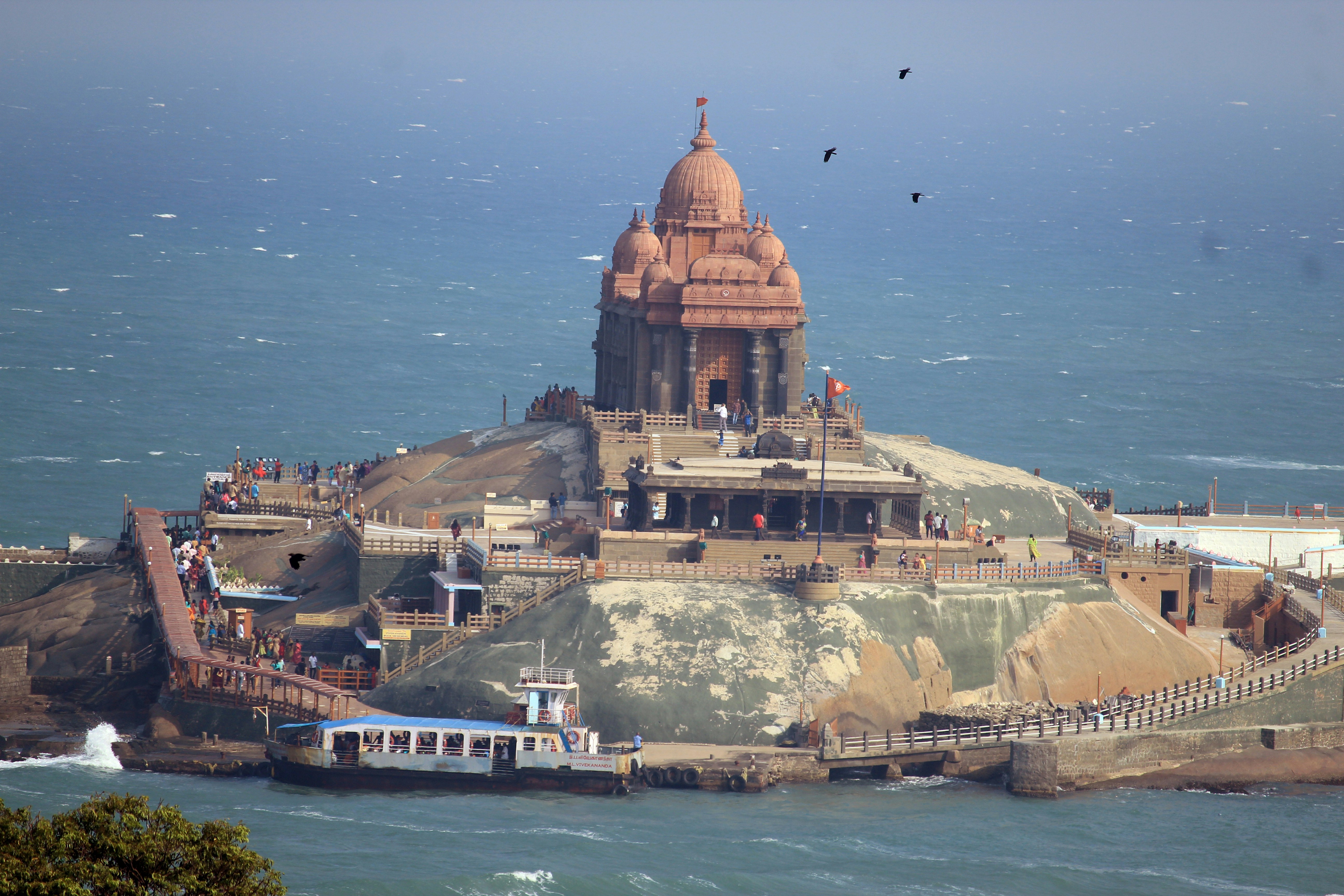 A coastal temple stands majestically on a rocky outcrop, surrounded by waves and visitors exploring the scenic site.