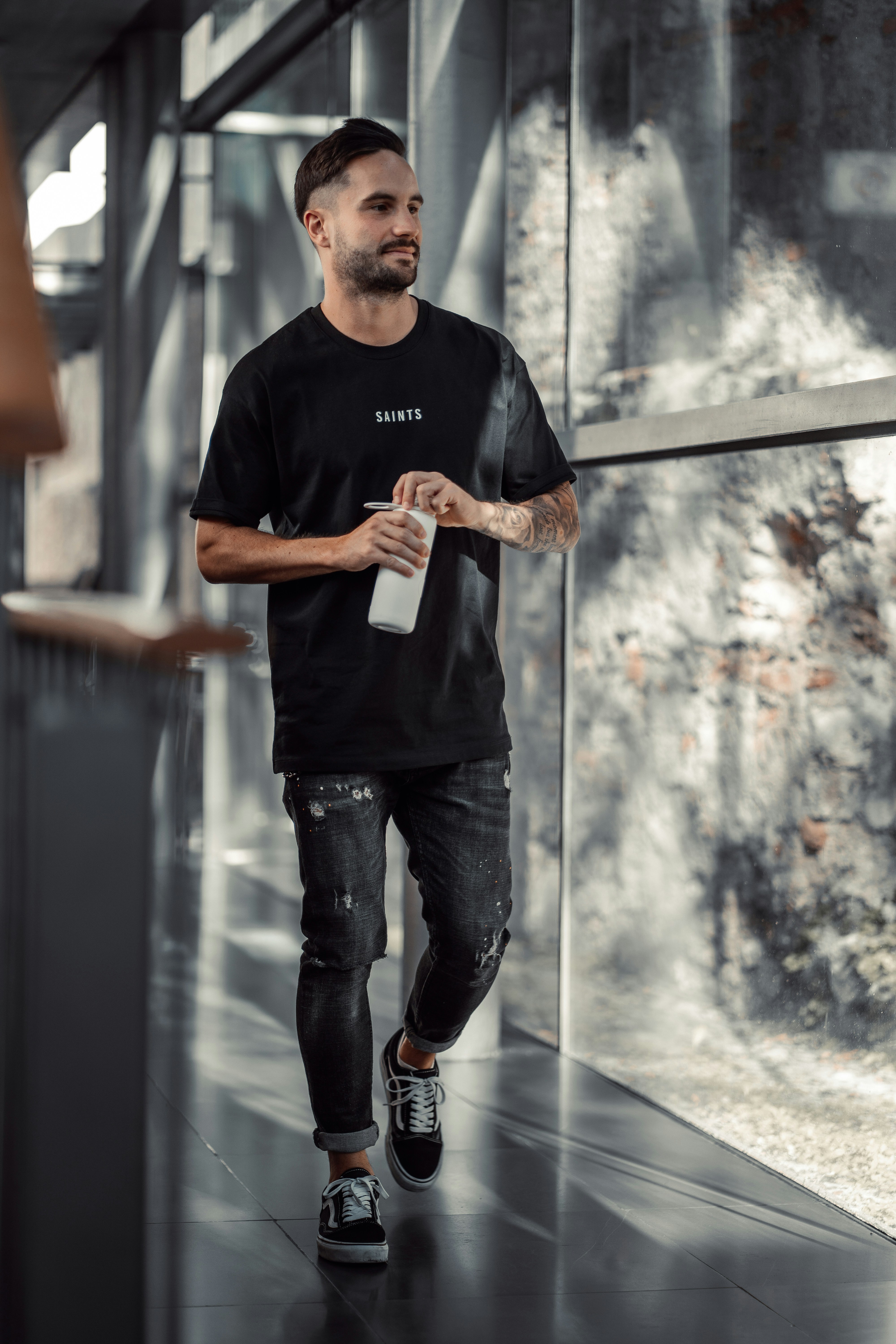 Man in a black t-shirt and distressed jeans walks through a modern interior, holding a drink. Sunlight streams through large windows, creating a dynamic play of light and shadow.