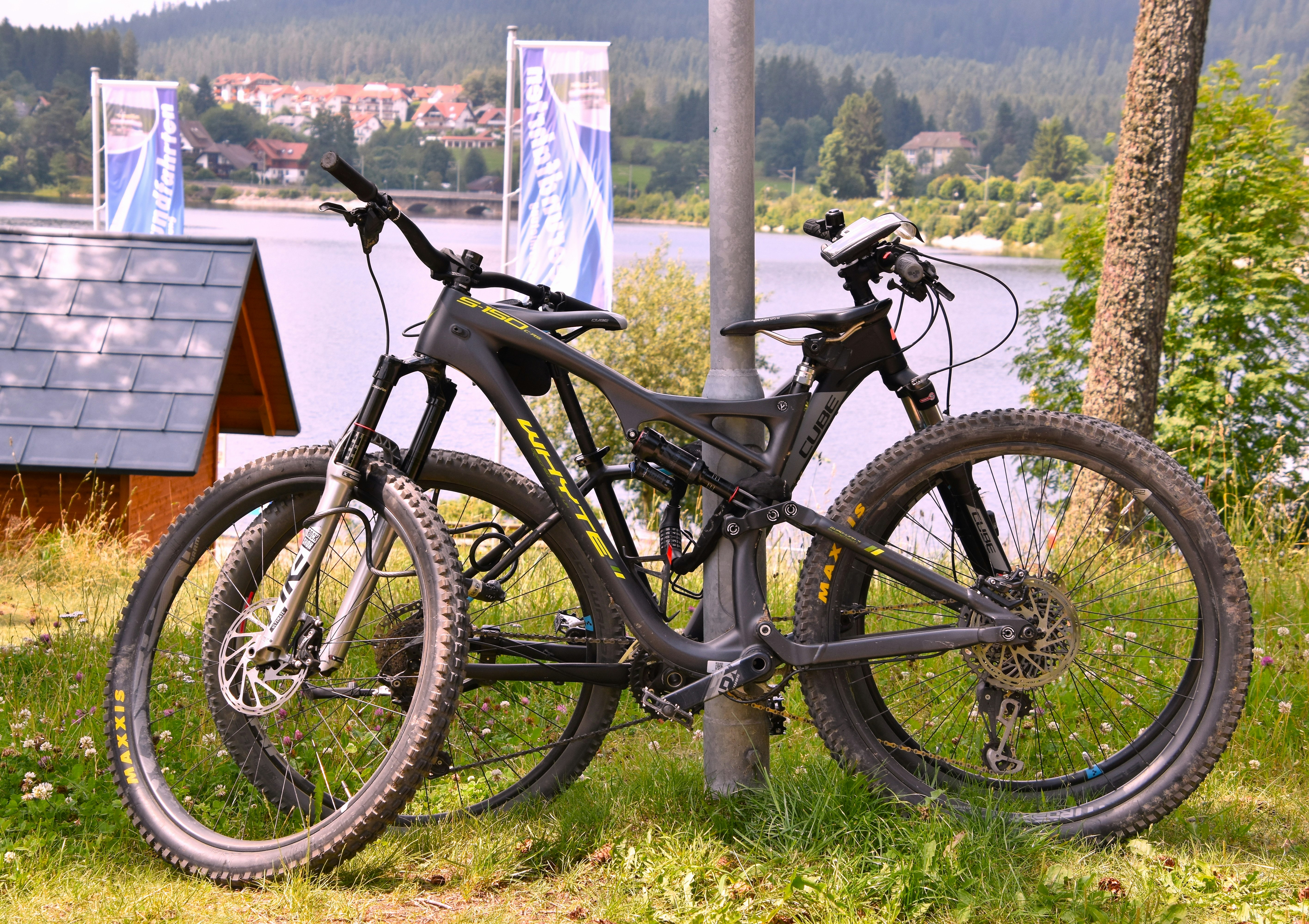 Mountain bike resting against a pole with a scenic lake and forest backdrop. The composition highlights the bike's rugged design and the serene environment.