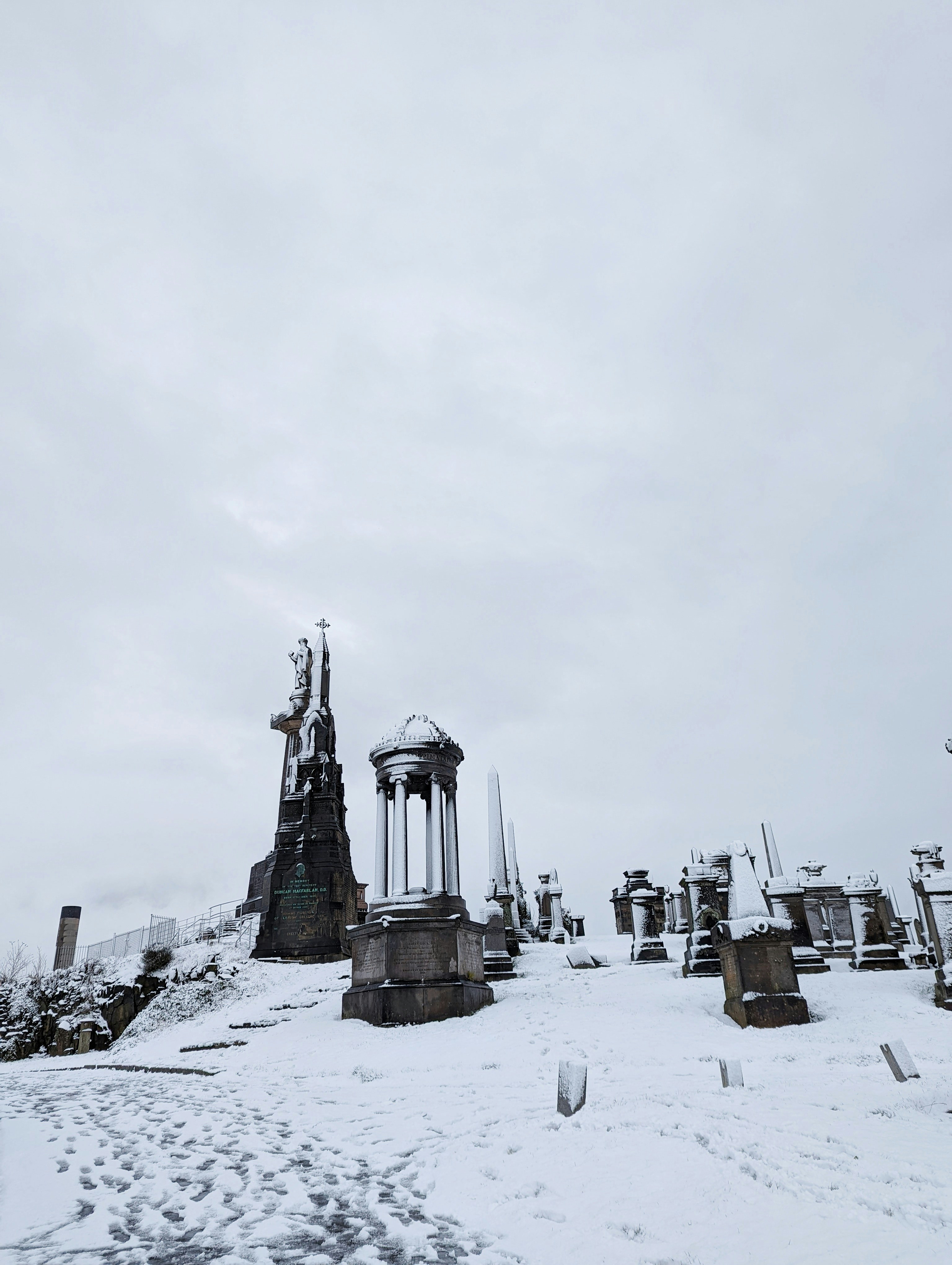 Snow-blanketed gravestones and monuments at Glasgow Necropolis under a gray sky.