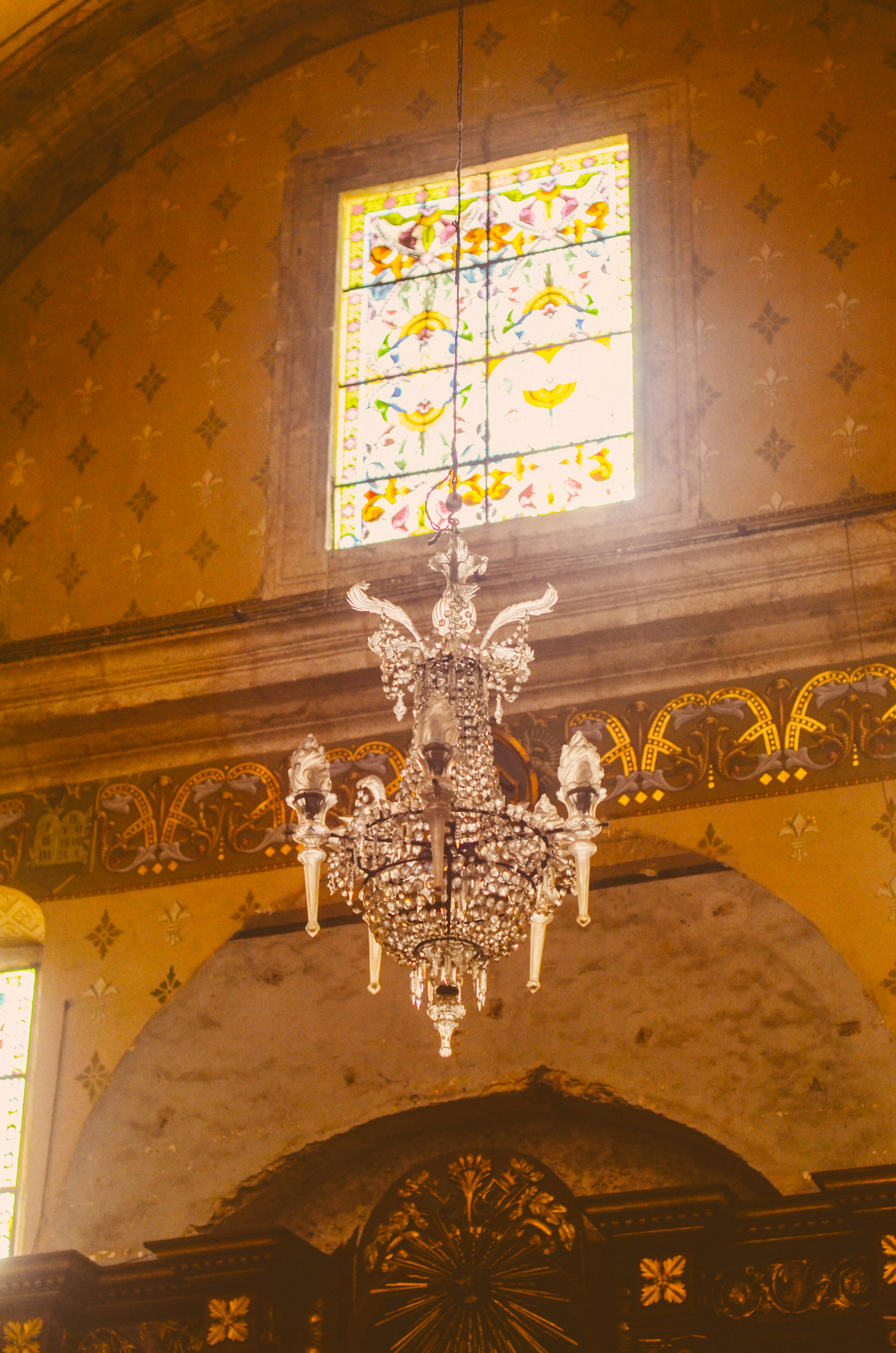 Intricately designed chandelier suspended beneath stained glass windows in a historic setting.