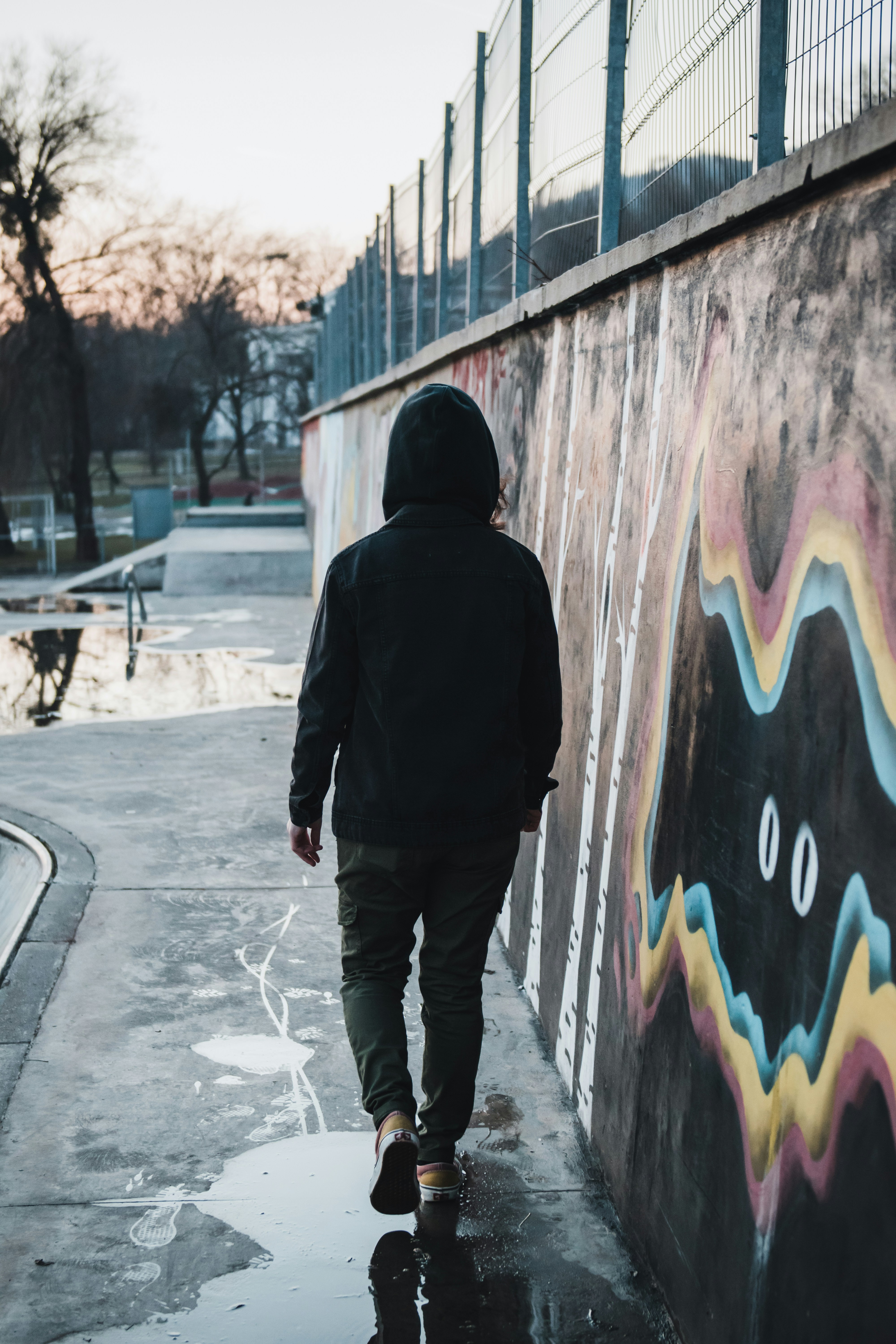 A lone figure walks along a graffiti-adorned path, mirroring the vibrant colors of the wall in nearby puddles. The scene captures a moment of introspection in an urban setting.