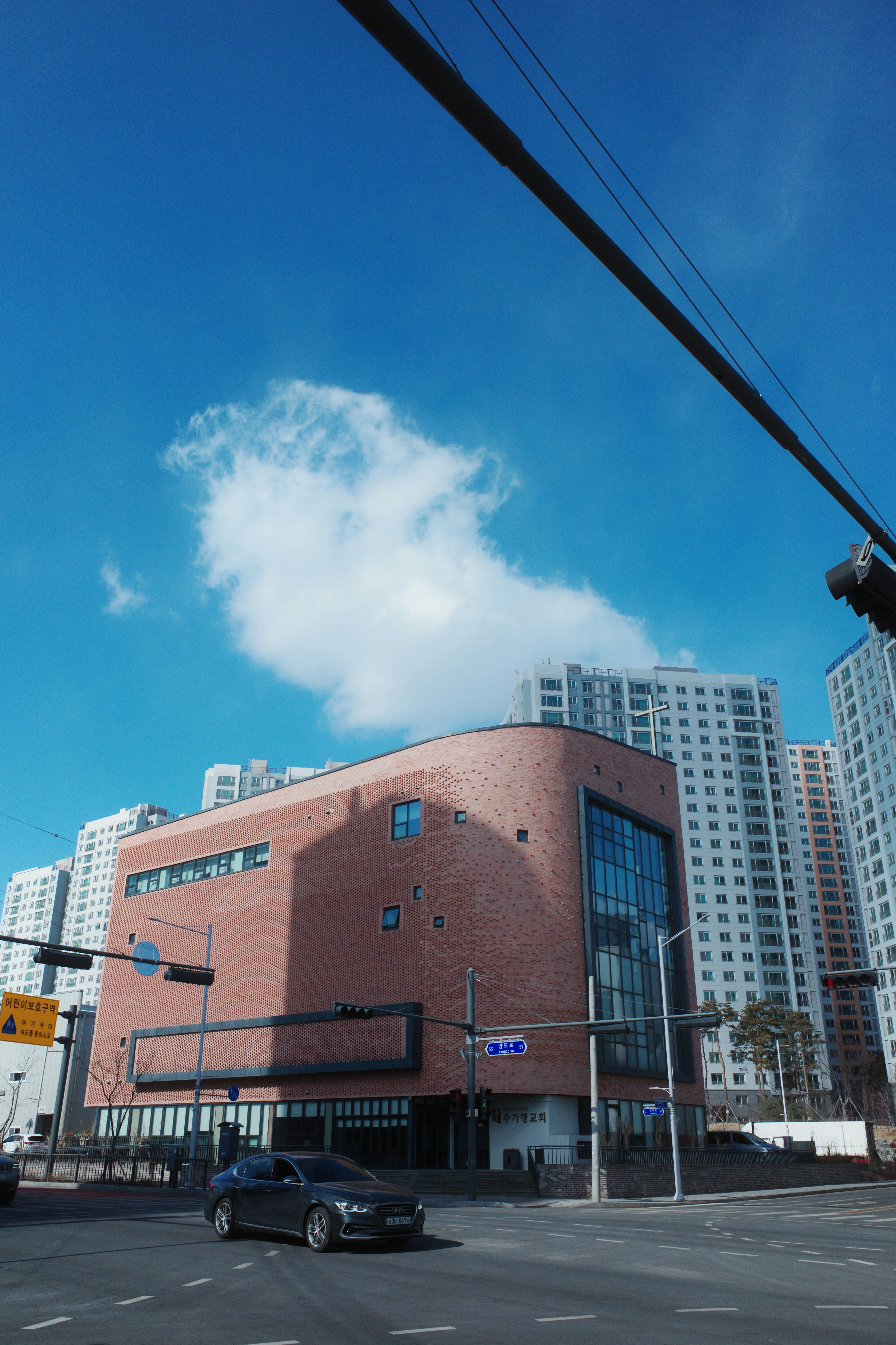 A contemporary brick building stands at a city intersection under a bright blue sky, with a single cloud floating above. The scene captures the blend of urban architecture and natural elements.