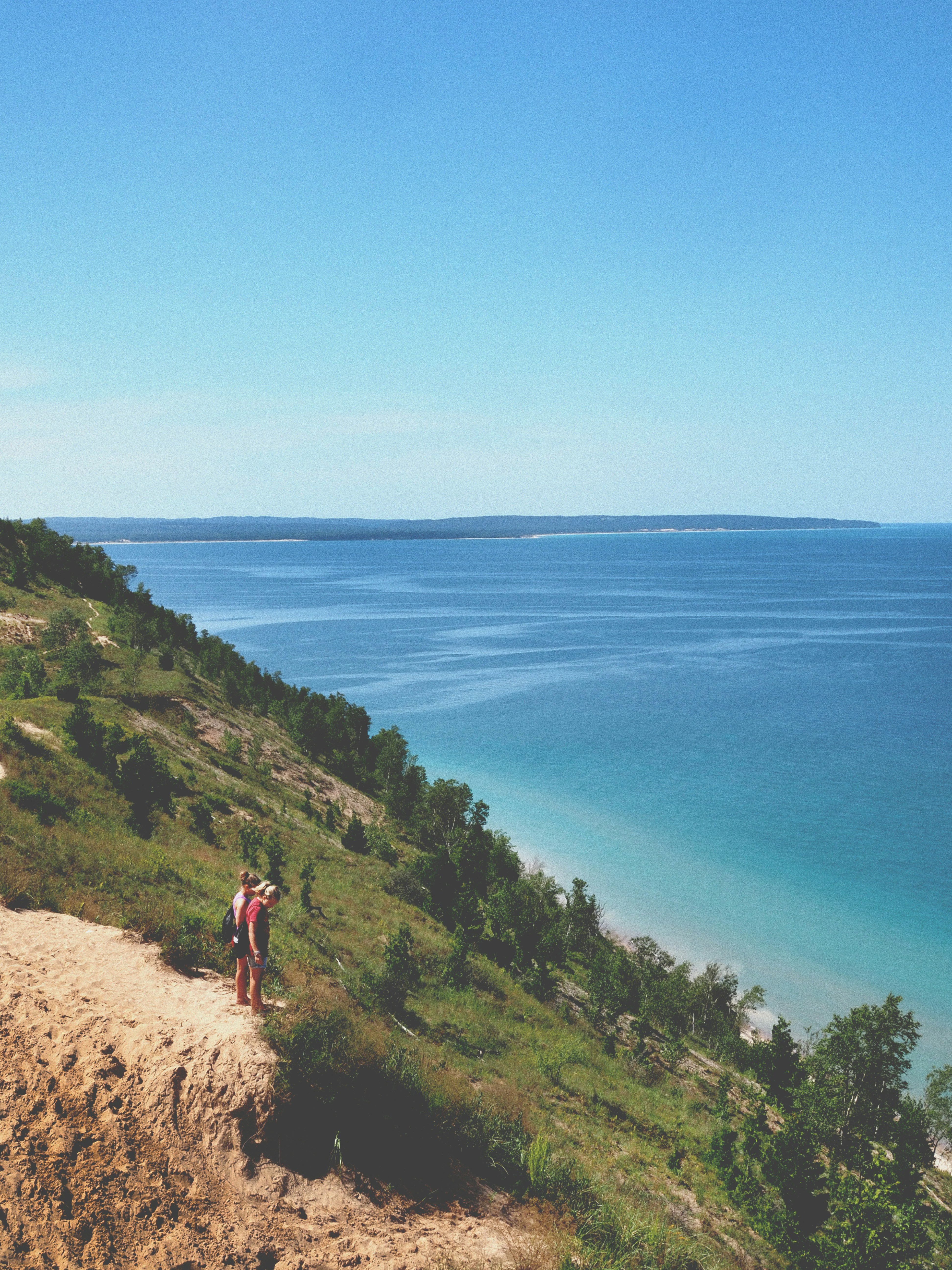 Two kids consider the mystery of the vastness of our planet as they stand on an ancient sand dune overlooking a great lake. | a couple of people standing on top of a lush green hillside